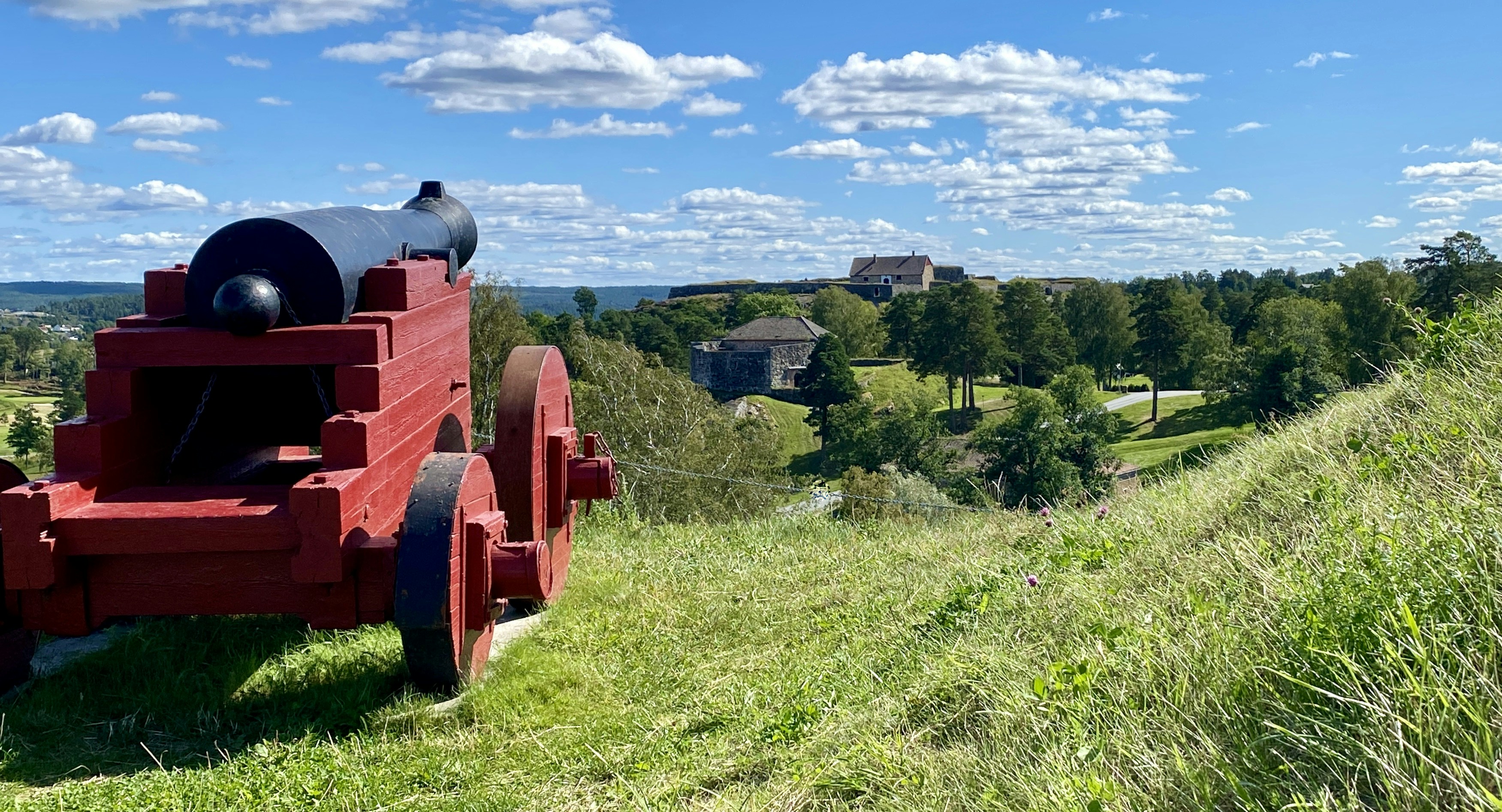 Old cannon at Fredriksten fortress in Halden, Norway. | Red cannon on grassy hill overlooking historic buildings
