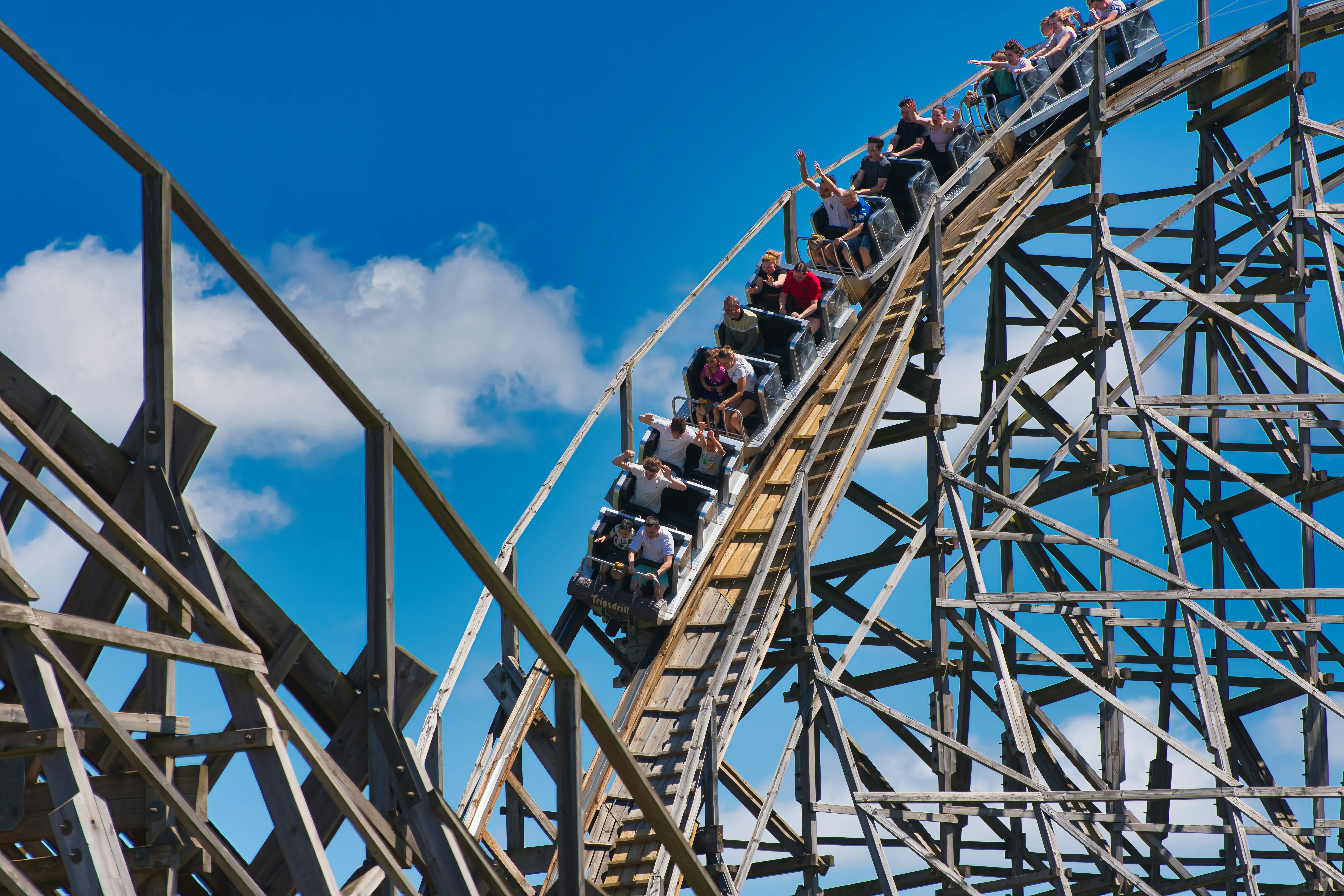 Riders experience the exhilarating climb of a wooden roller coaster against a bright blue sky with fluffy clouds.