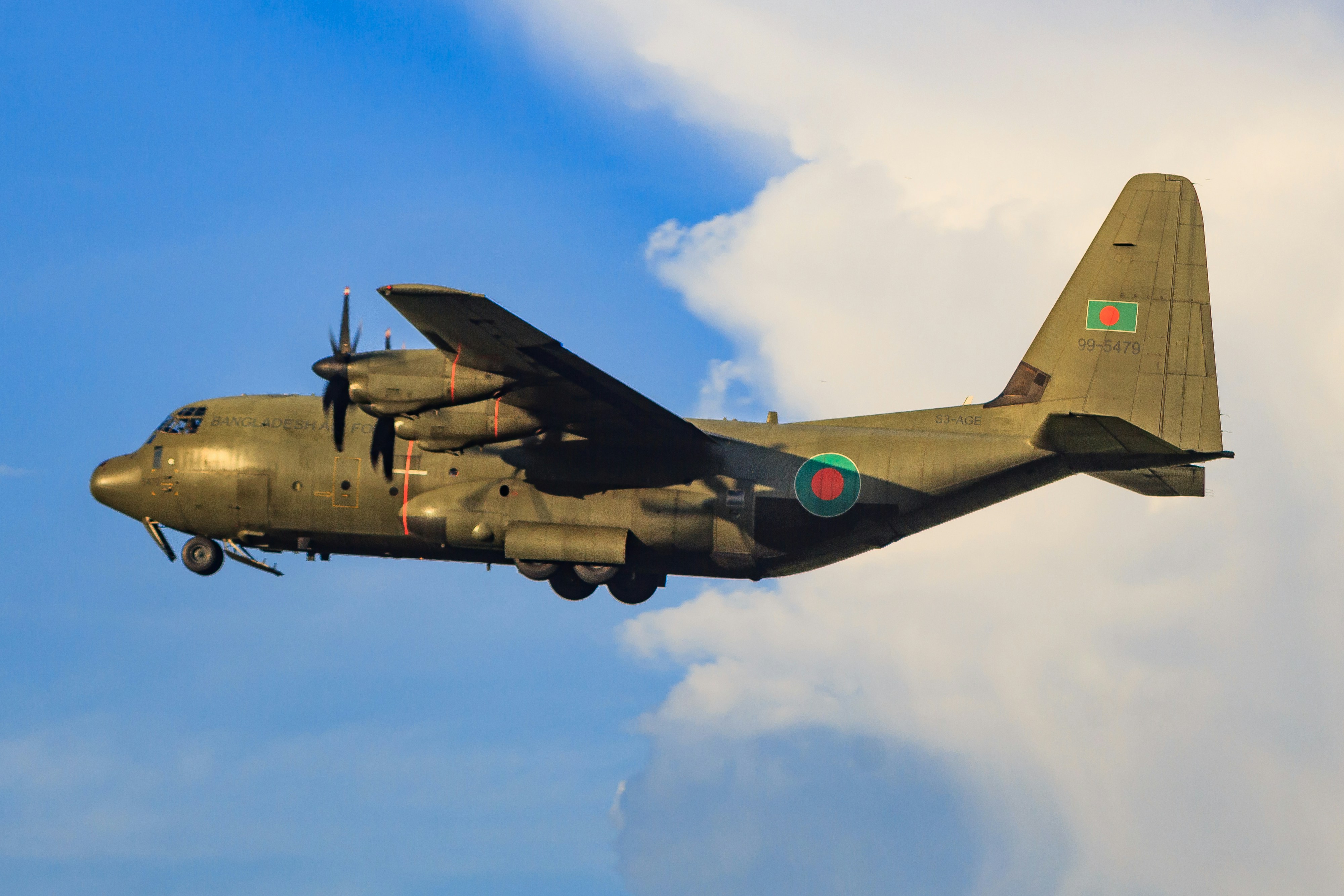 Military transport aircraft flying against a backdrop of fluffy clouds. The aircraft features the national insignia of Bangladesh.