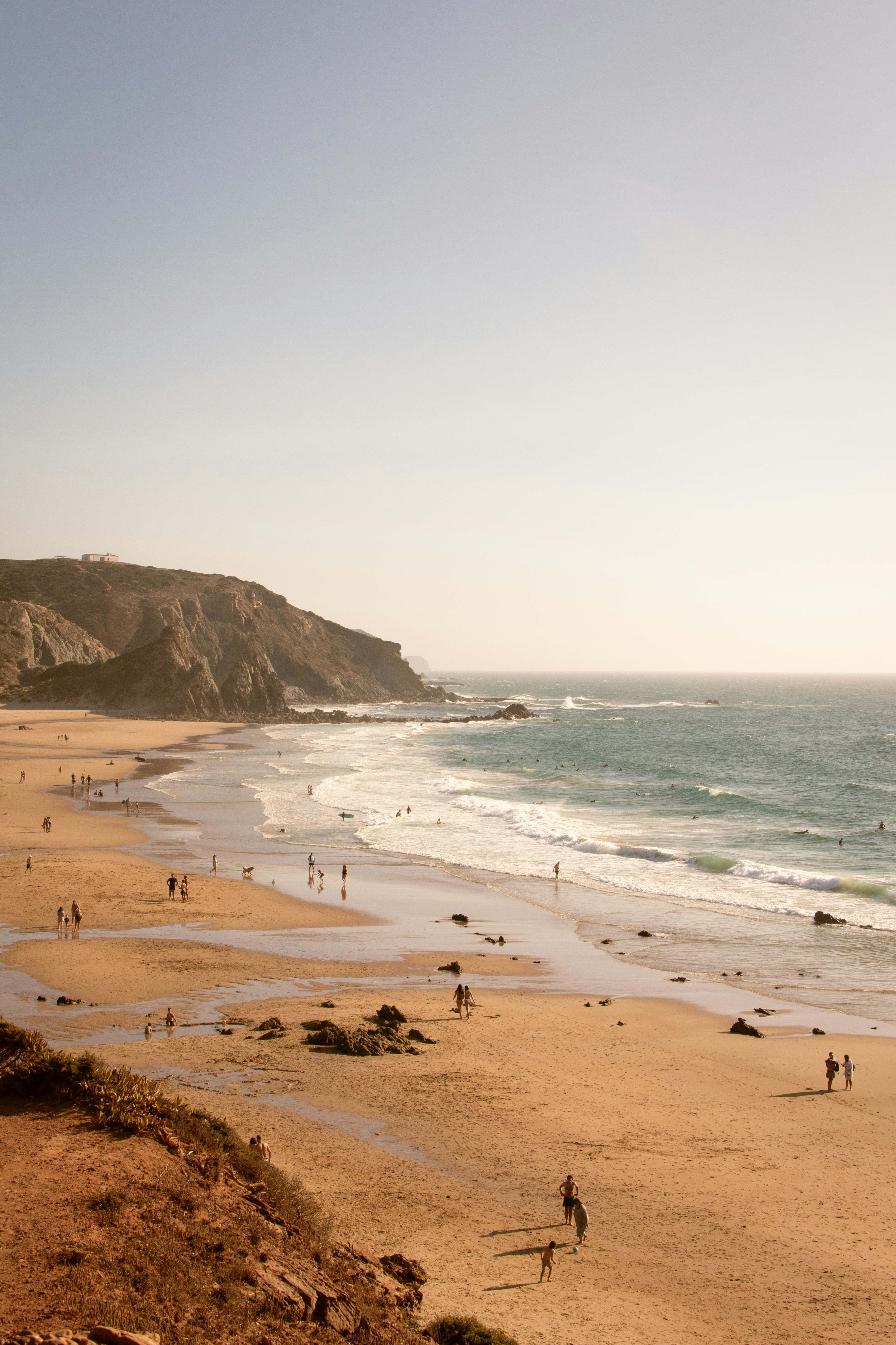 Golden sands stretch along the coastline, dotted with beachgoers enjoying the serene waves and rocky cliffs in the background.