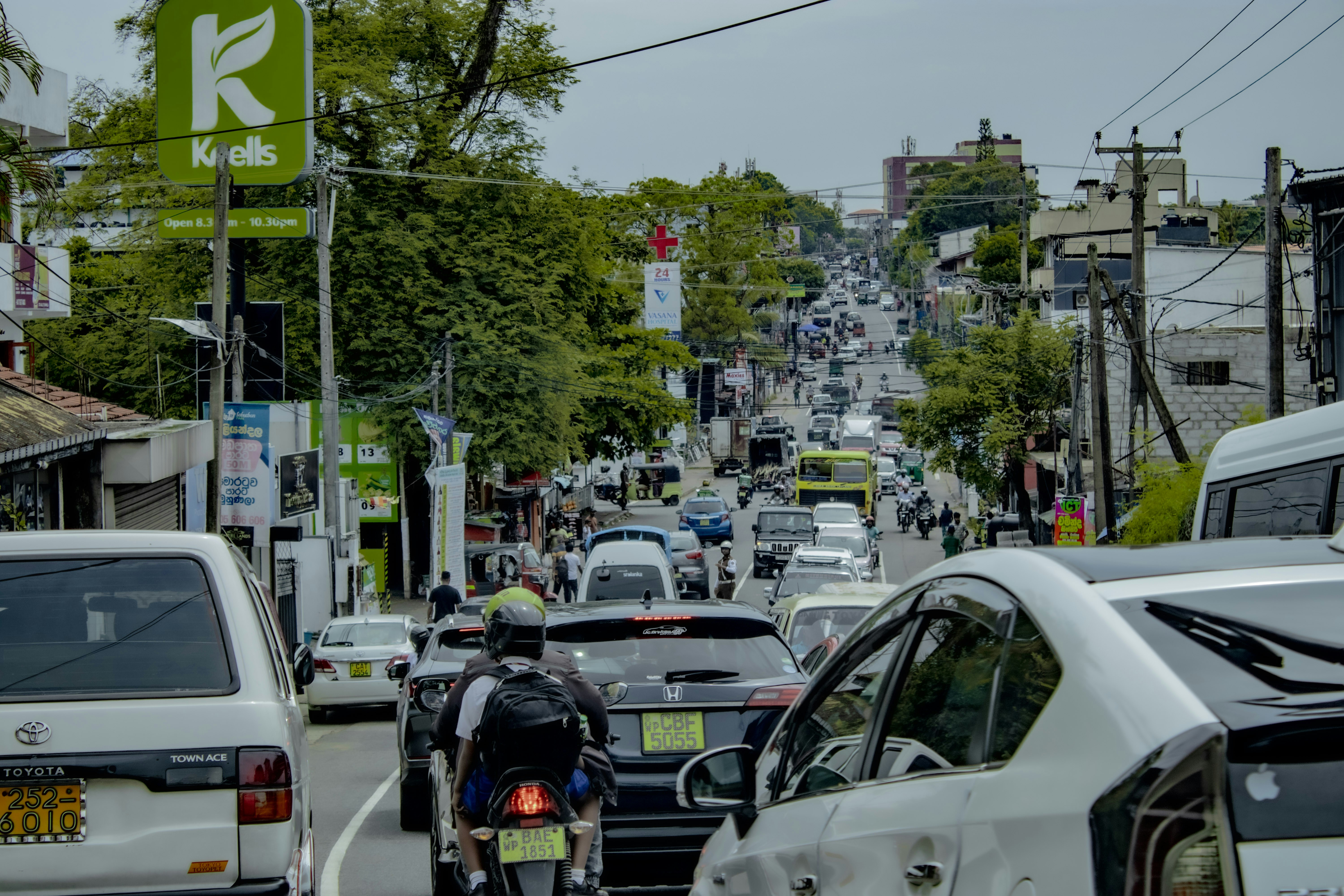 Busy street scene showcasing a blend of vehicles and pedestrians amidst urban greenery. A prominent sign adds character to the bustling atmosphere.