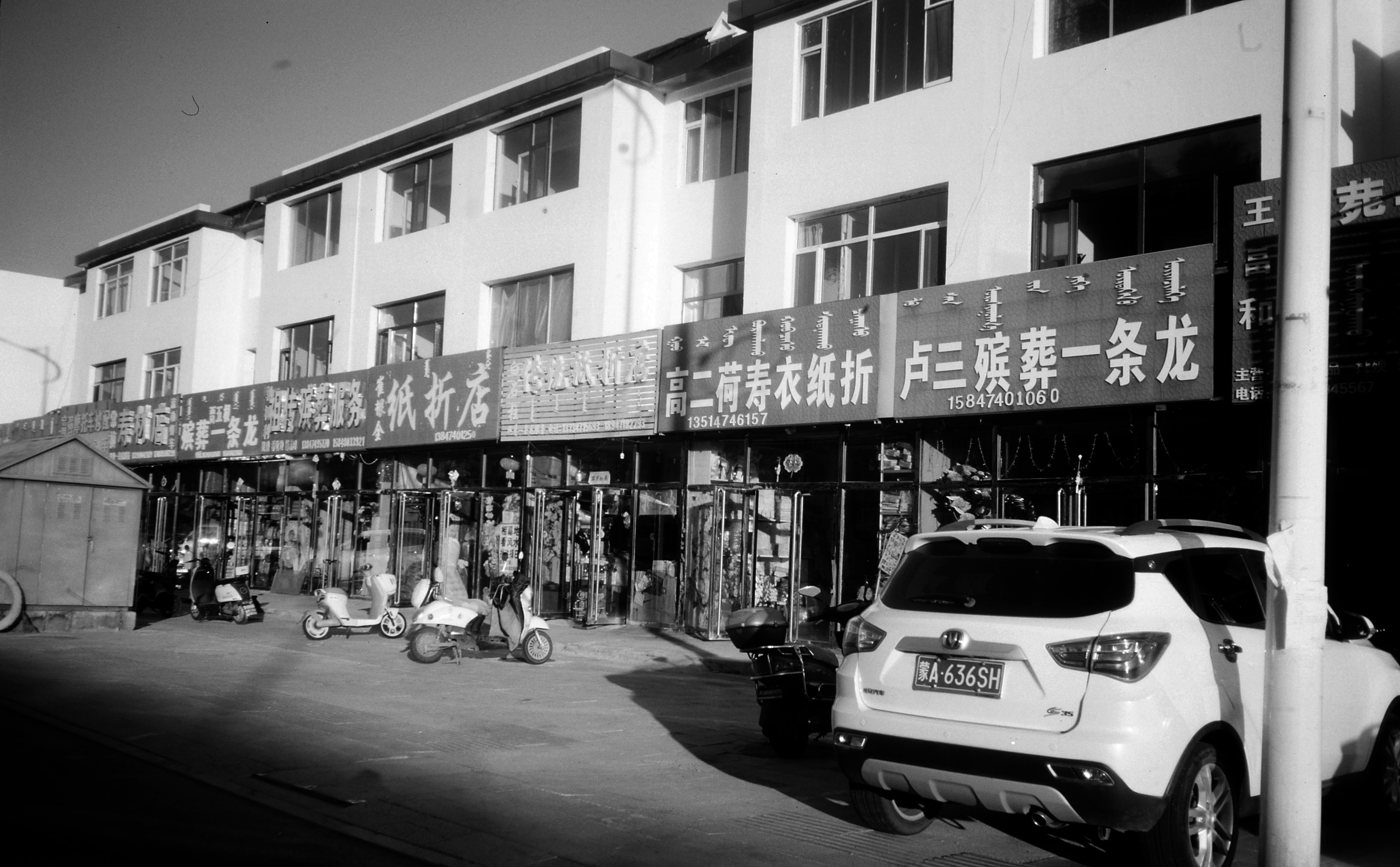 The population of my hometown has been declining for years. Most young people have left to seek job opportunities elsewhere, leaving behind many elderly residents. As a result, there are now many funeral supply stores in the town. | Street scene with shops, vehicles, and signage.