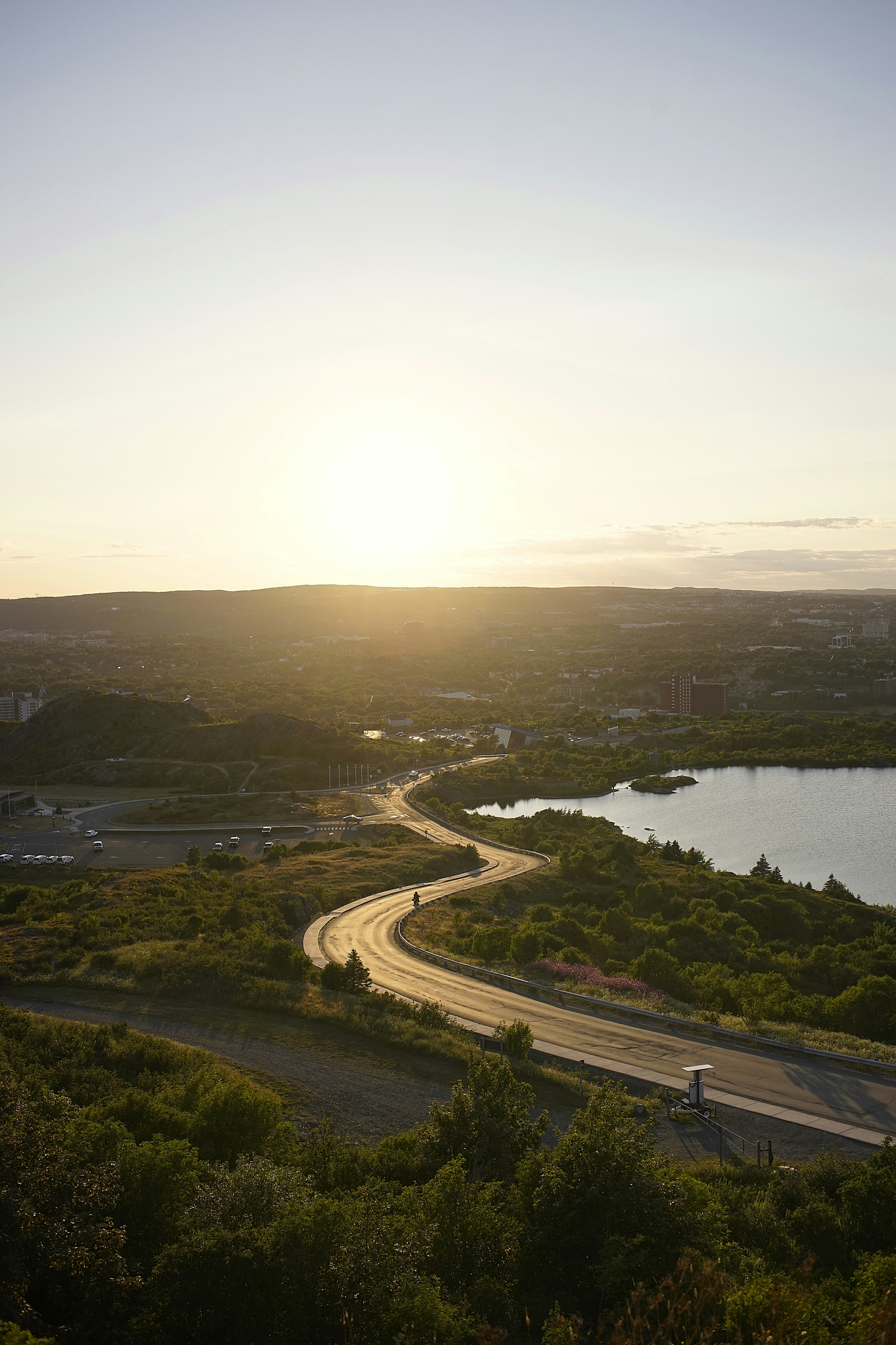 Curved road meandering through lush terrain under a setting sun, with a serene lake reflecting the twilight hues.