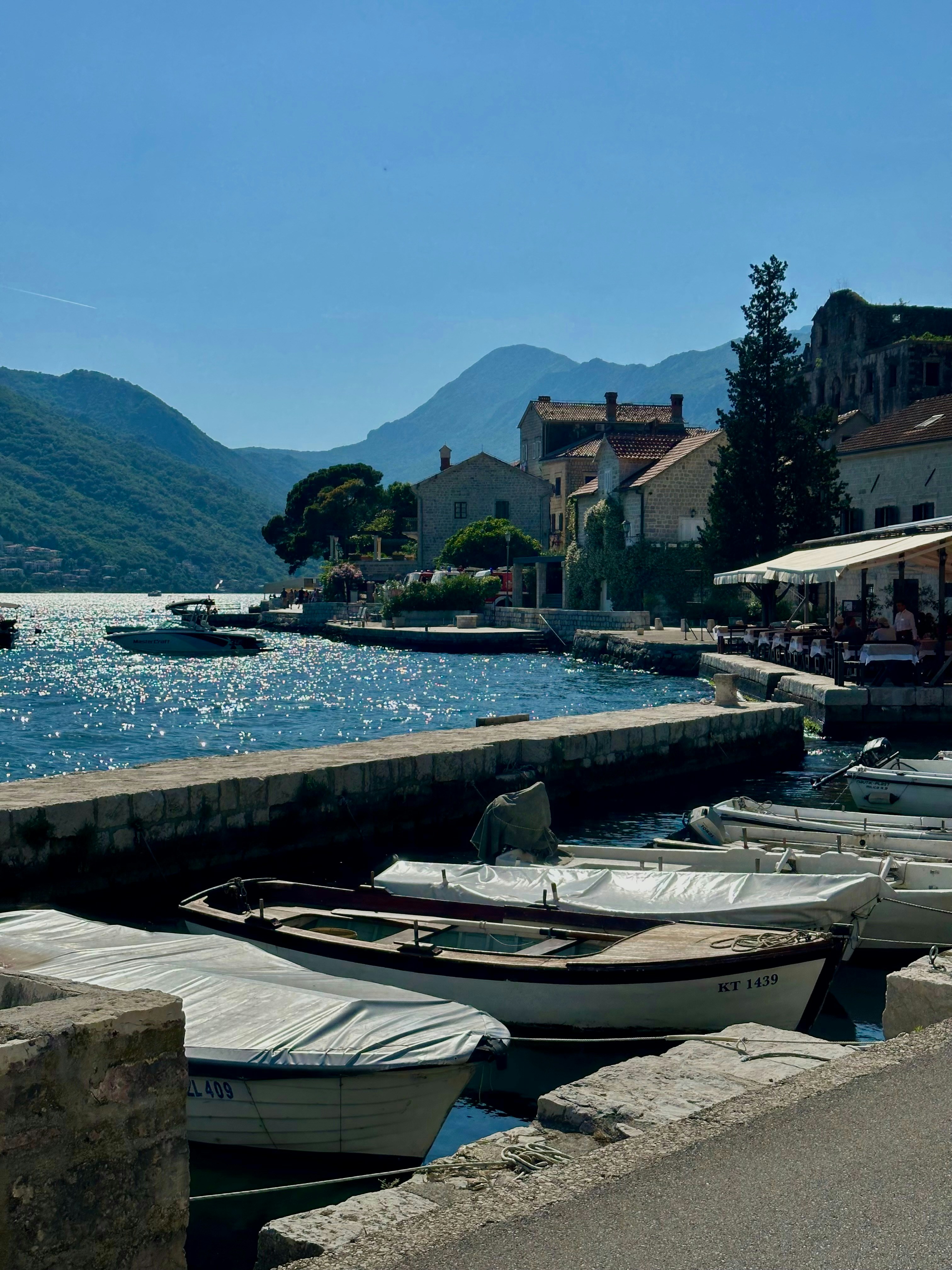Charming harbor scene featuring boats docked along a sunlit waterfront with picturesque buildings and mountains in the background.