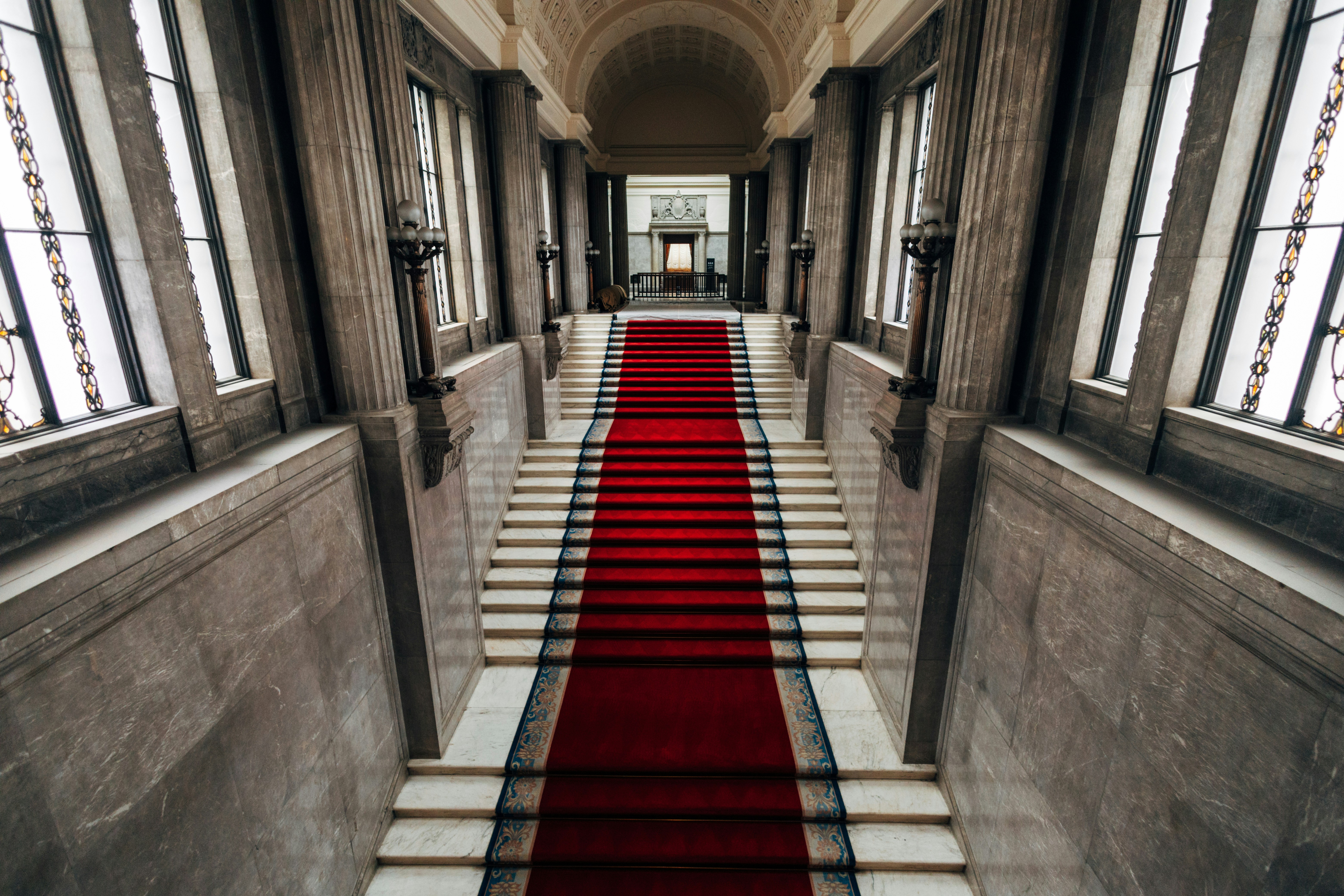 Luxurious red carpet leading down a grand staircase in a historic building, flanked by elegant windows and ornate architecture.