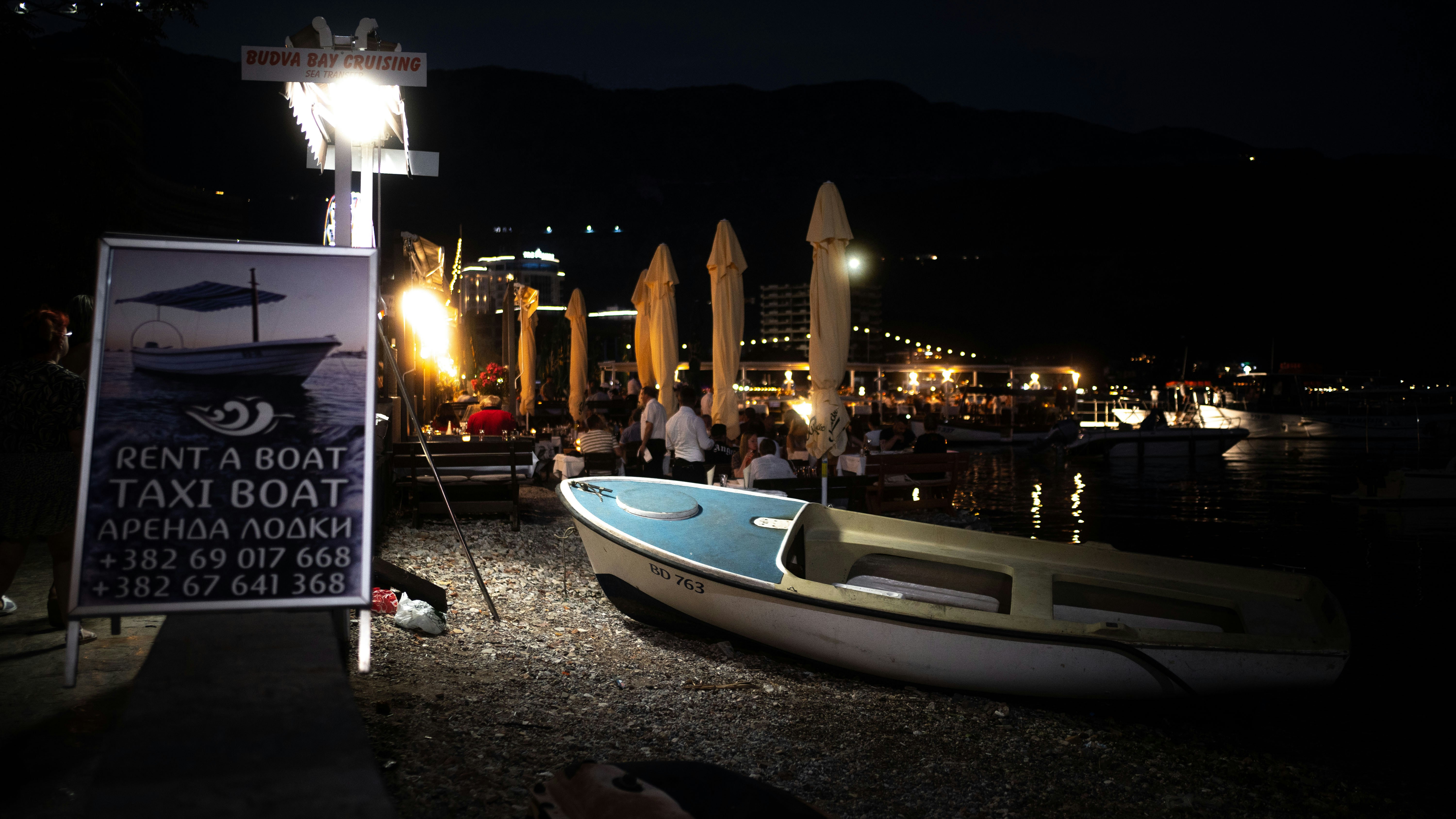 A small taxi boat rests on the shore at night, illuminated by the glow of seaside restaurants and marina lights. Captured in Budva, Montenegro, this scene reflects the vibrant nightlife, coastal charm, and travel atmosphere of the Adriatic Sea. | Boats on a beach near a restaurant at night