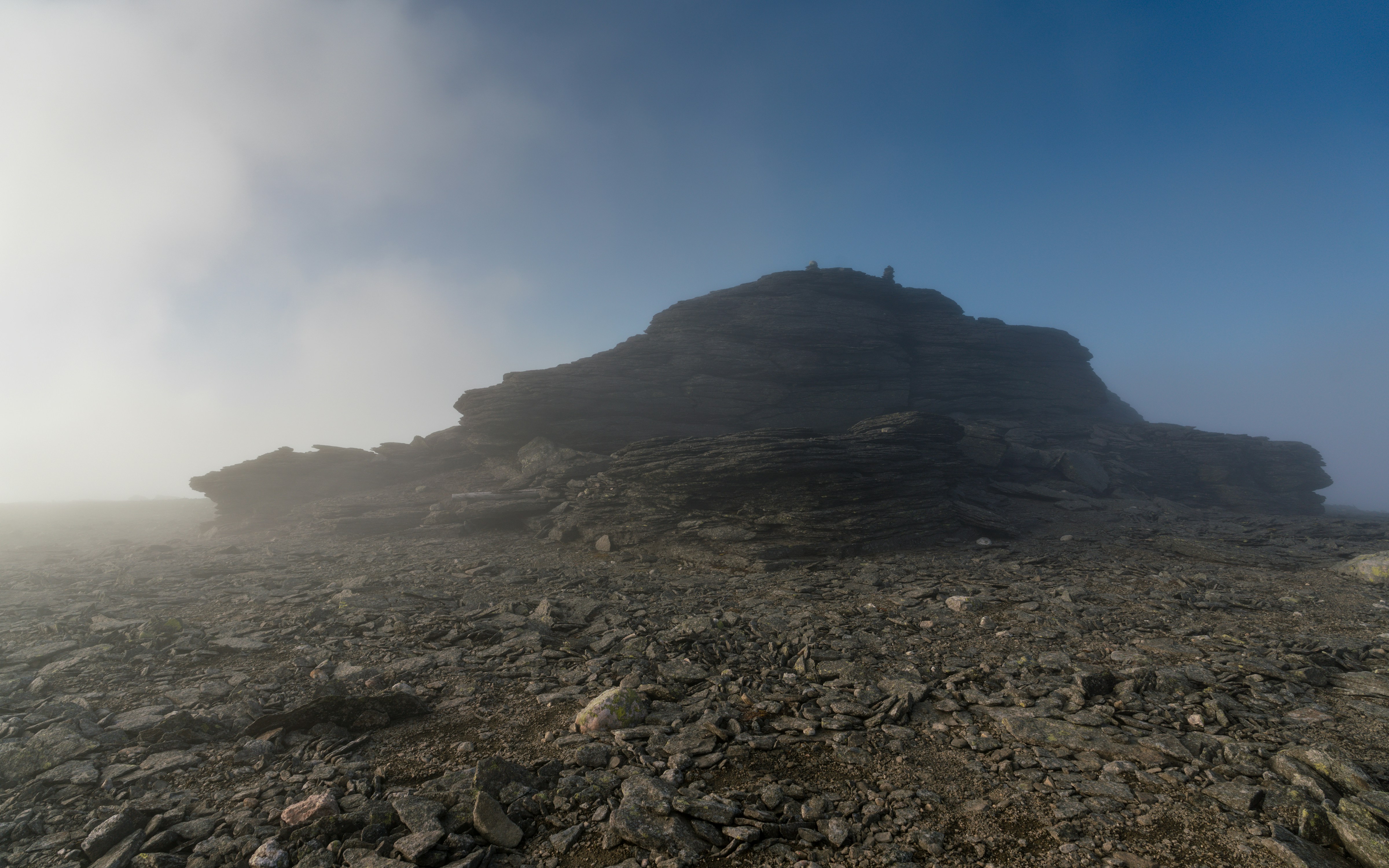 Mysterious rocky formation partially obscured by fog, creating an ethereal atmosphere. The rugged terrain suggests an ancient landscape shaped by time.