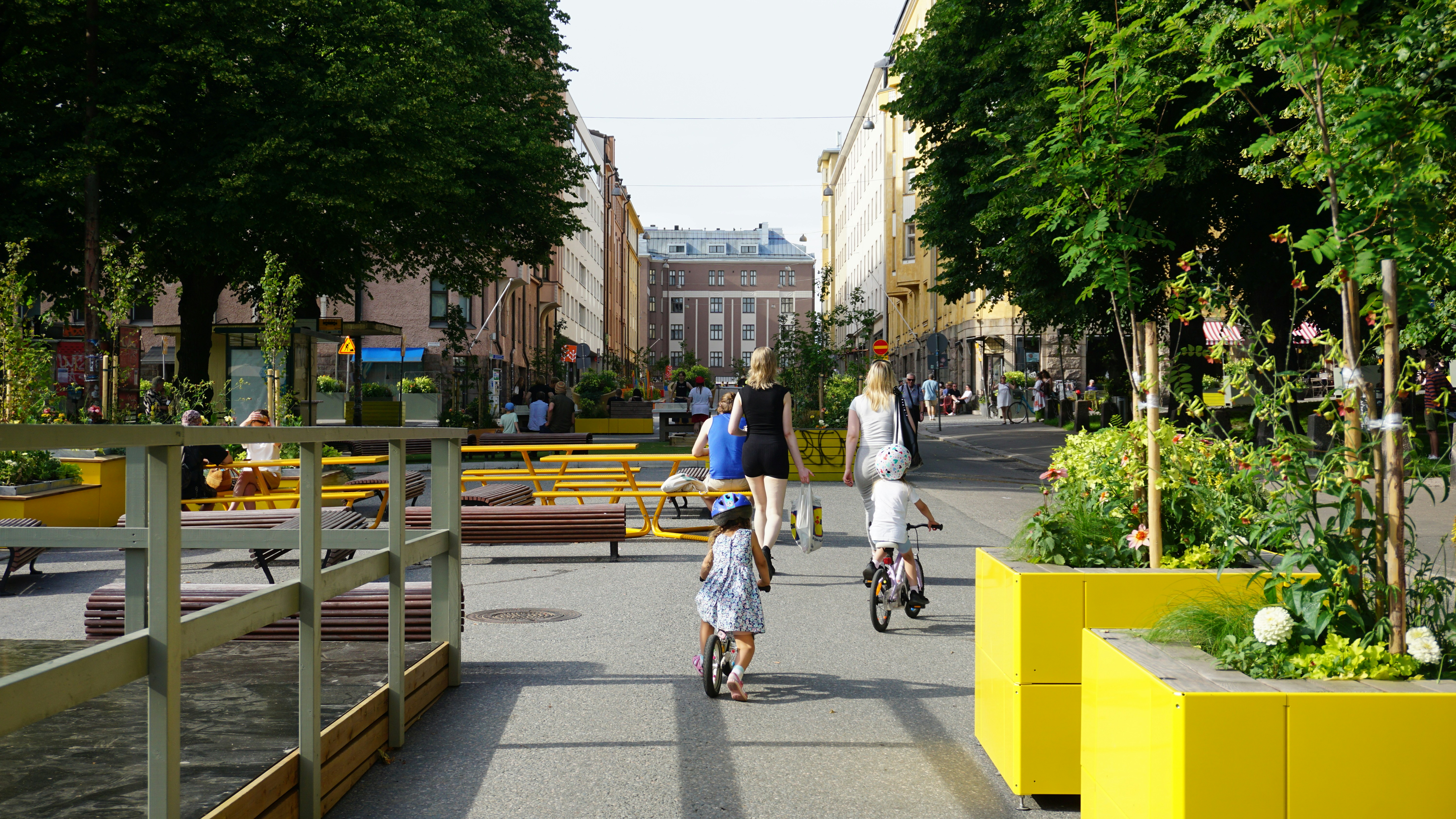 Two children riding scooters along a vibrant urban pathway, flanked by lush greenery and colorful planters. The scene captures the essence of community and outdoor leisure.
