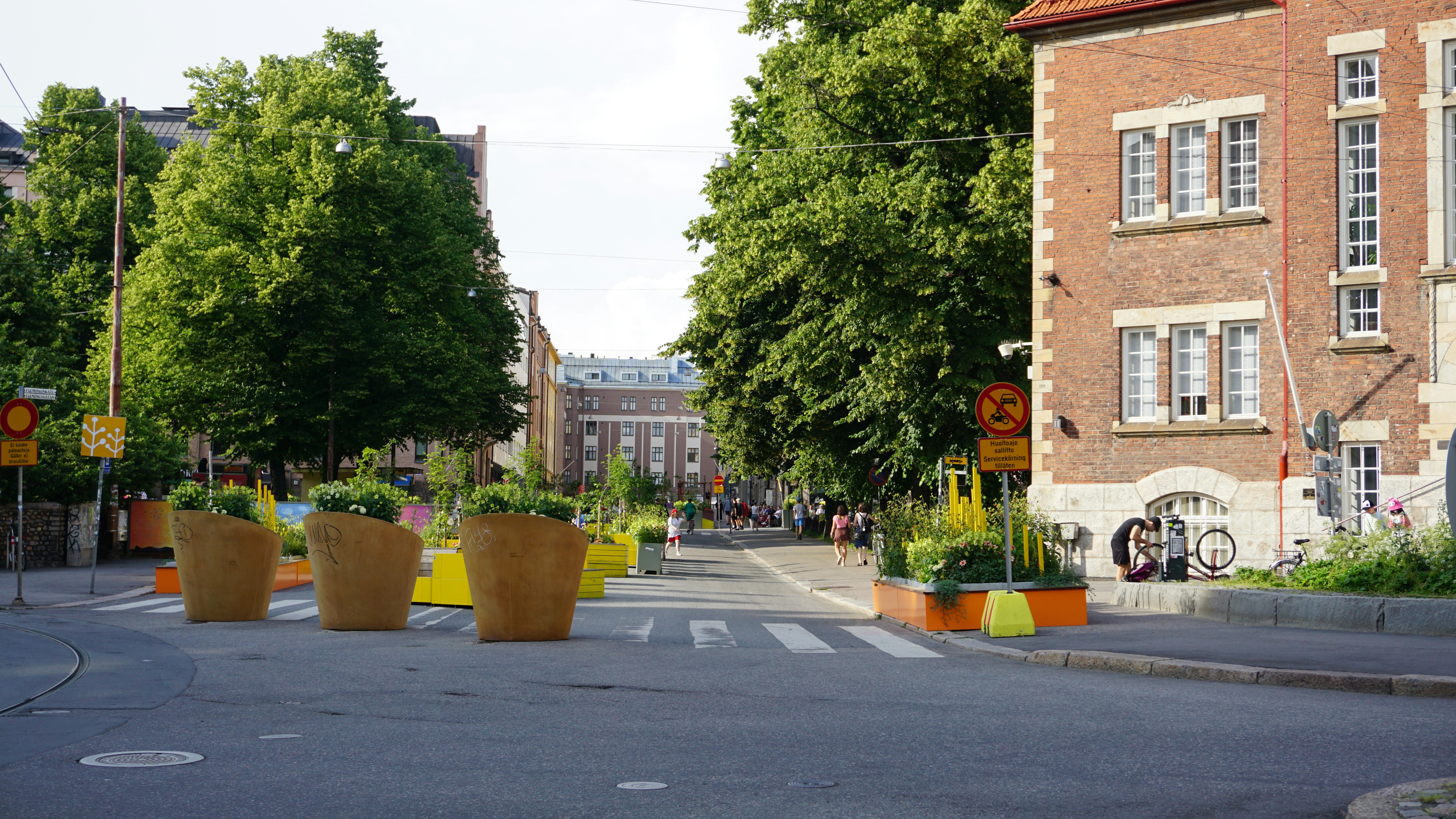 Colorful planters line a pedestrian-friendly street, creating a vibrant urban environment. People stroll in the background, highlighting community engagement.