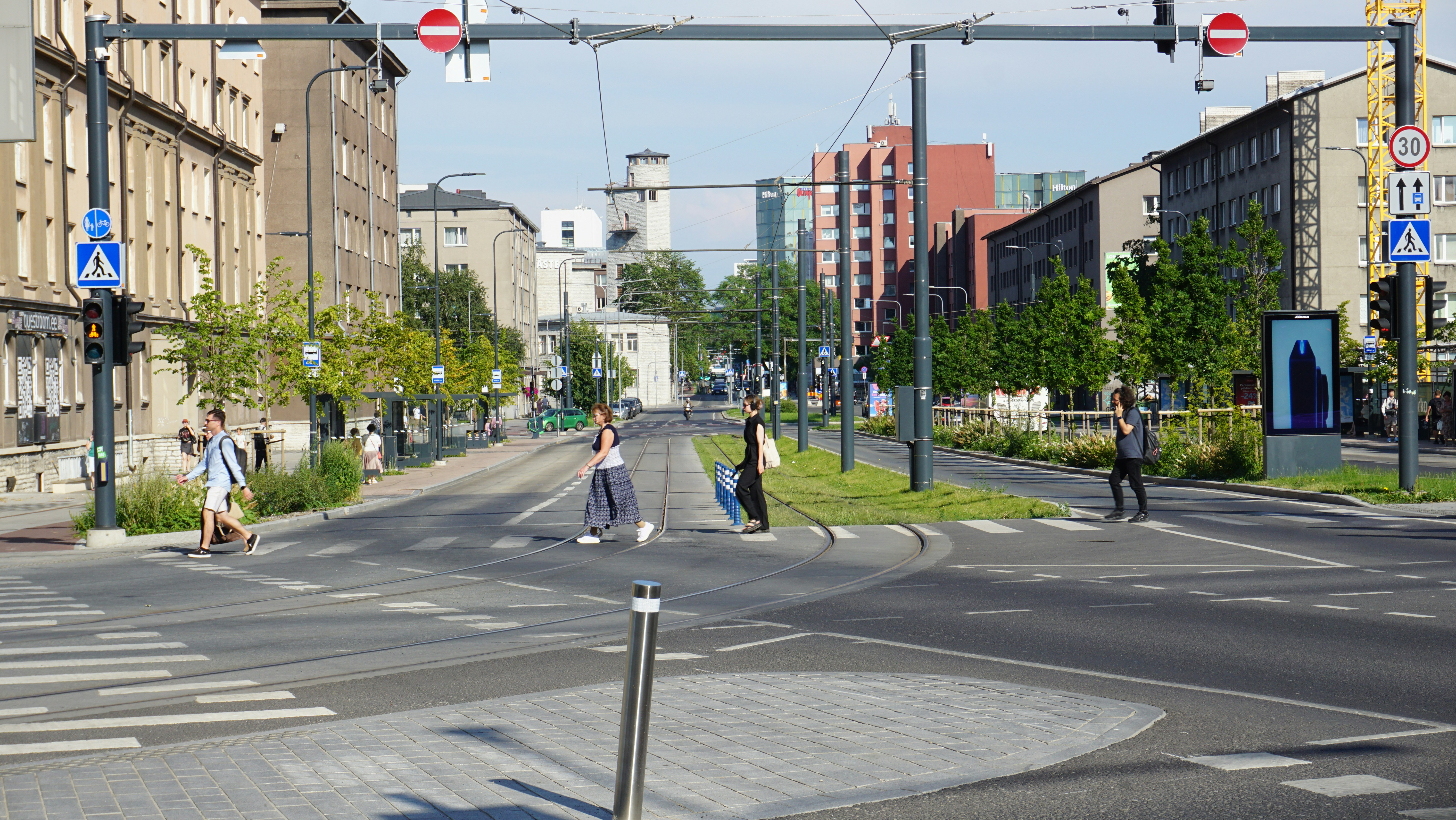 Pedestrians crossing a busy intersection in an urban landscape, with modern buildings and greenery framing the scene.