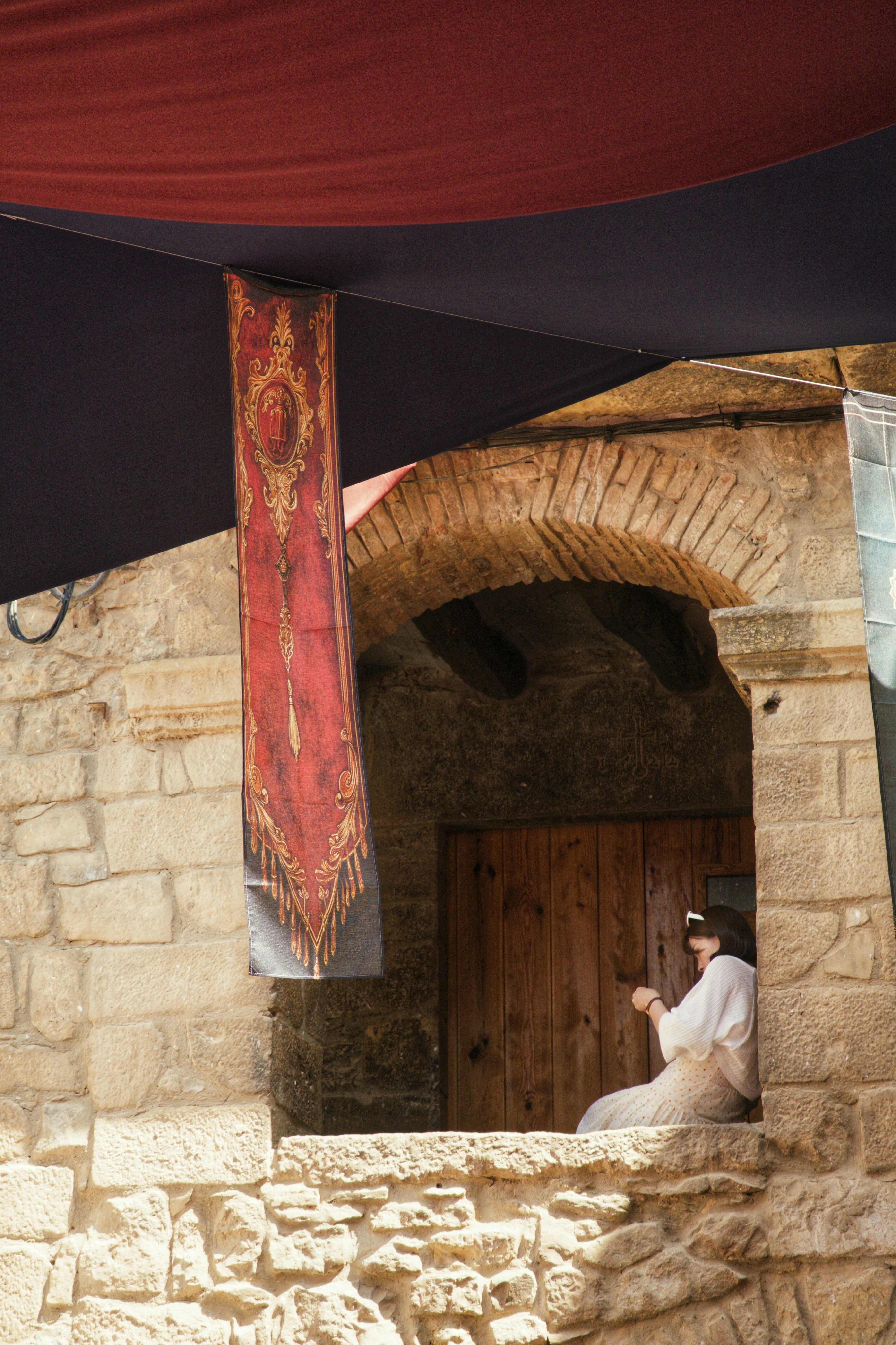 A woman in period attire sits quietly in a stone archway, framed by colorful banners and rustic architecture. The scene evokes a sense of history and contemplation.