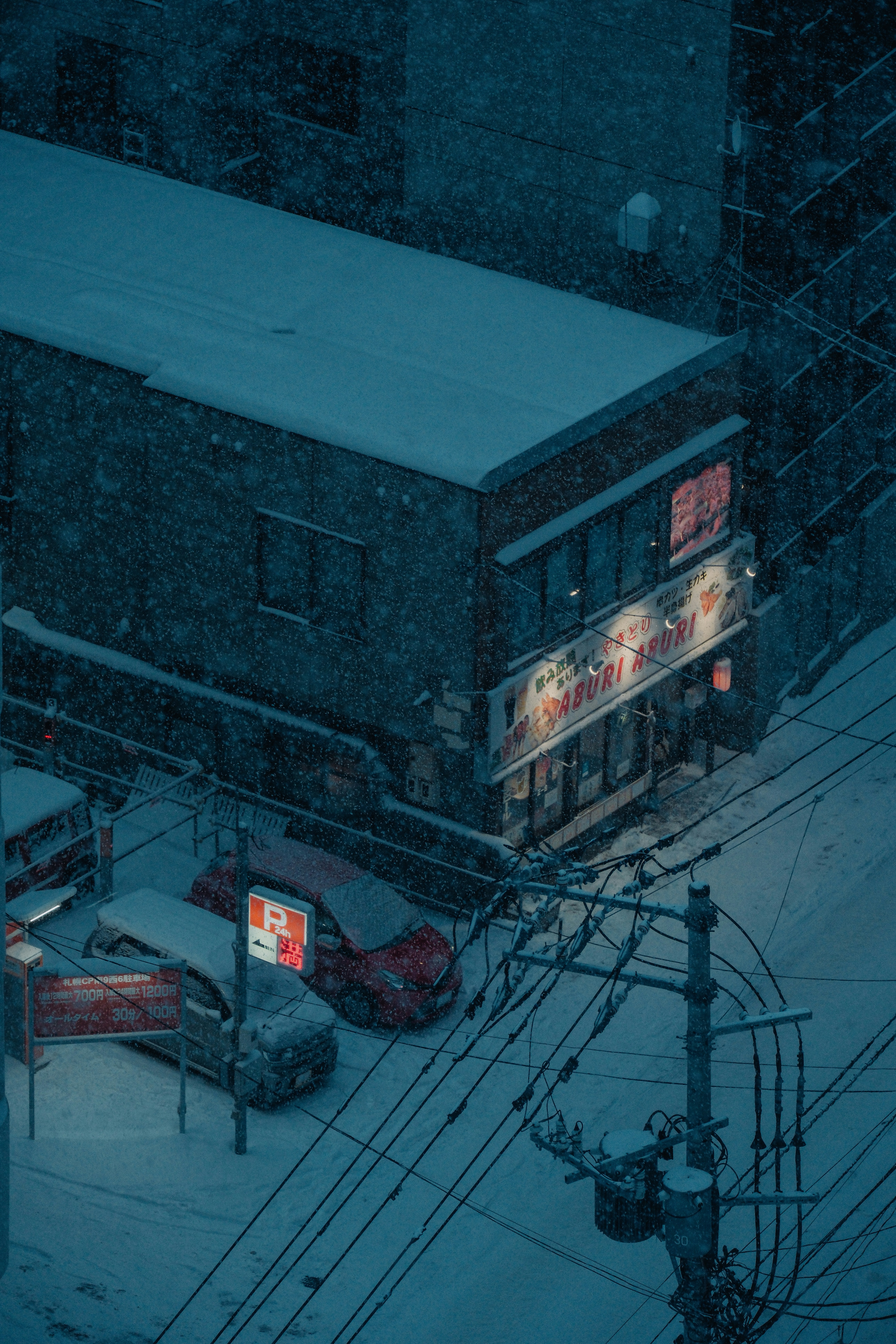 A cozy restaurant illuminated by warm lights amidst a heavy snowfall, with cars blanketed in snow and power lines crisscrossing the scene.