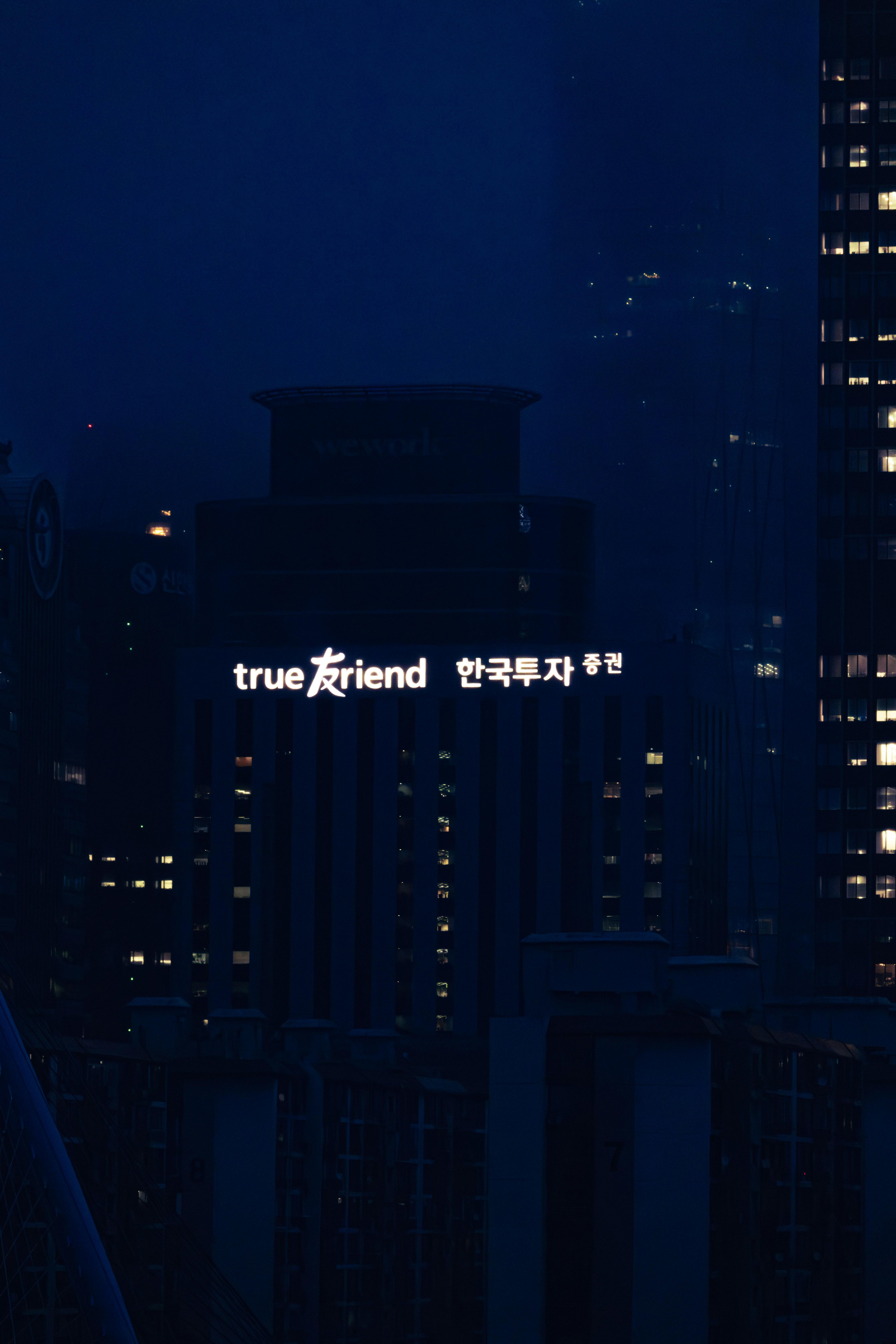 A tall office building glows with illuminated signage at night, surrounded by dark skyscrapers and scattered window lights in a moody cityscape. | City skyline at night with illuminated buildings