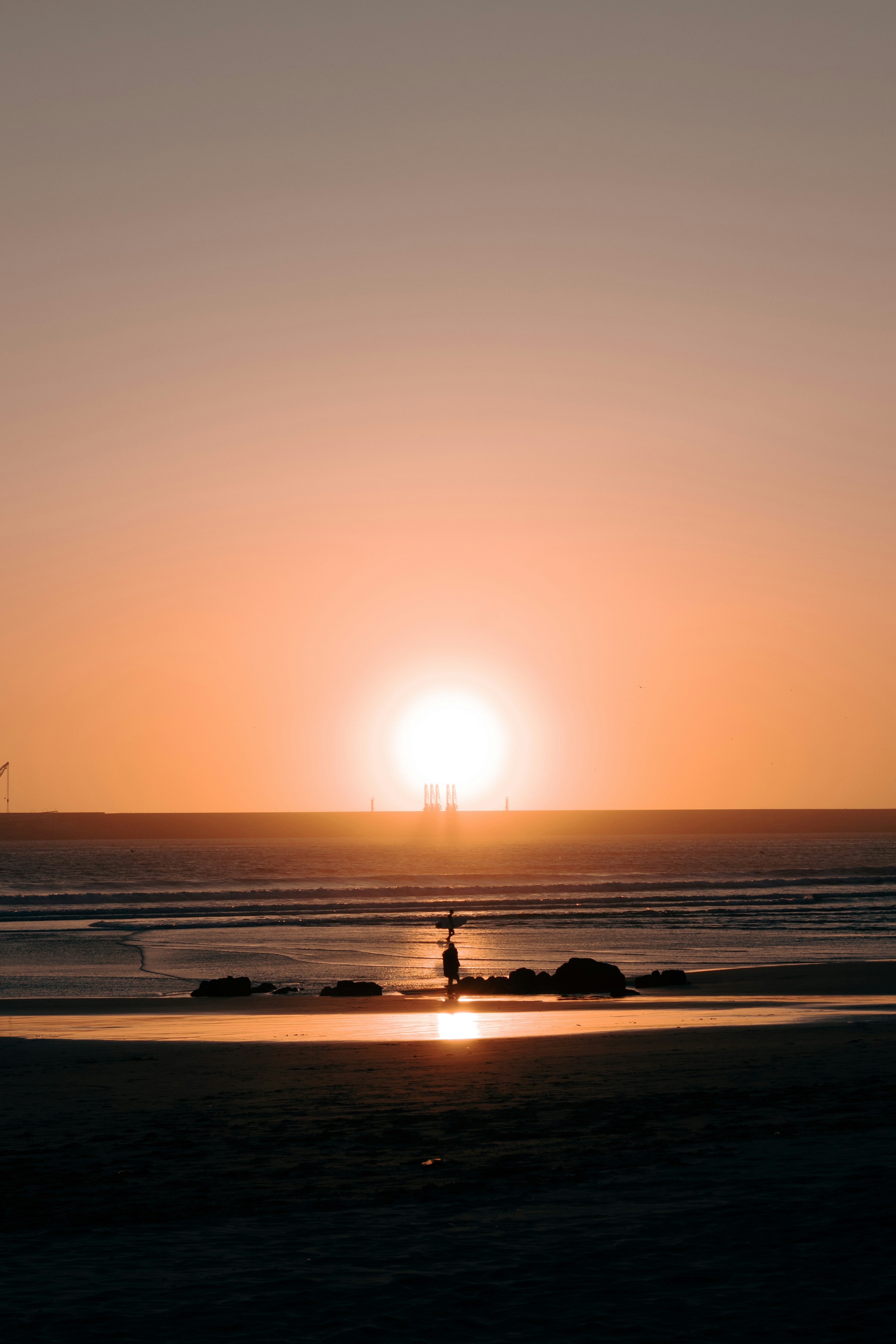 A lone figure stands on the beach, silhouetted against a vibrant sunset, with a distant horizon featuring wind turbines. The serene scene captures the essence of tranquility.