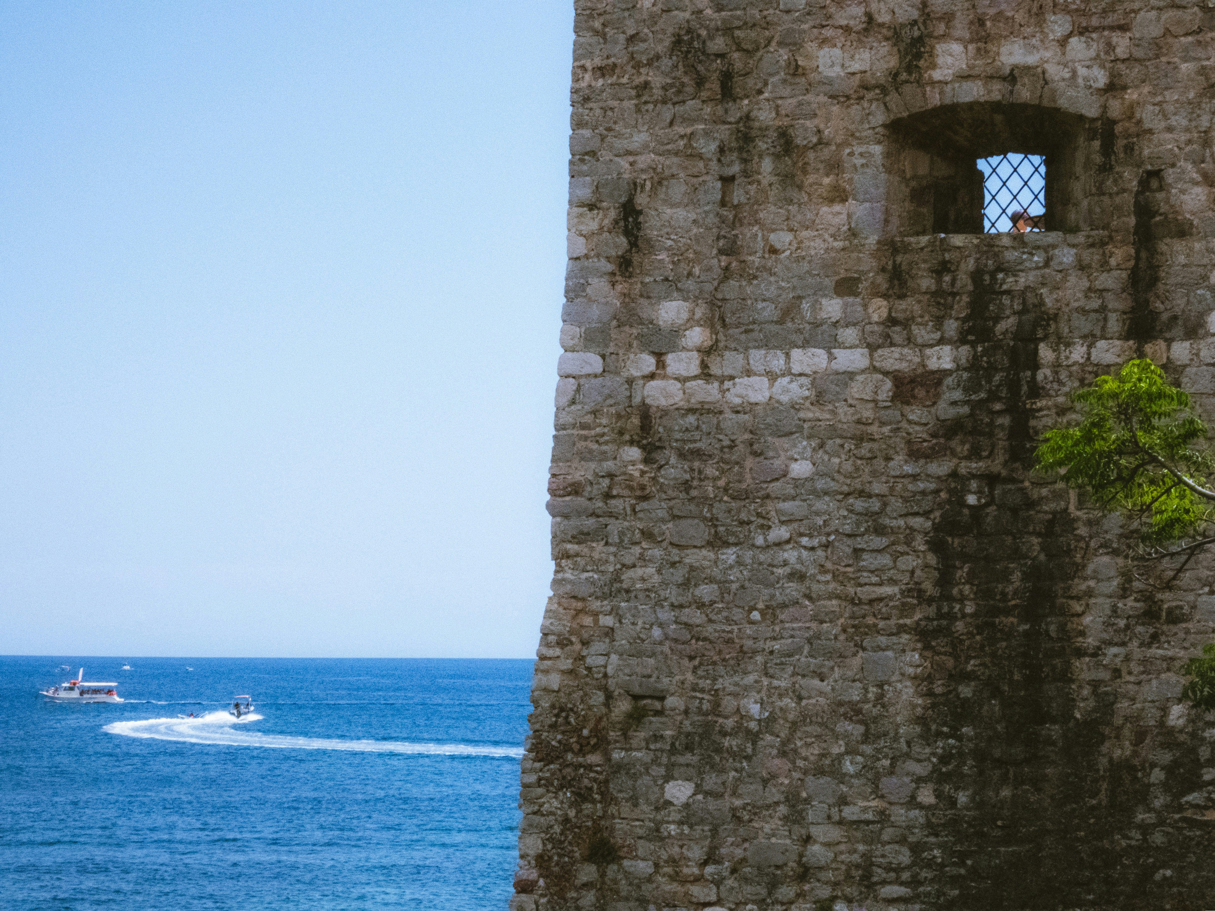 Old stone fortress wall with a small window overlooking the blue sea and passing boats. | Stone tower overlooks ocean with boats sailing.