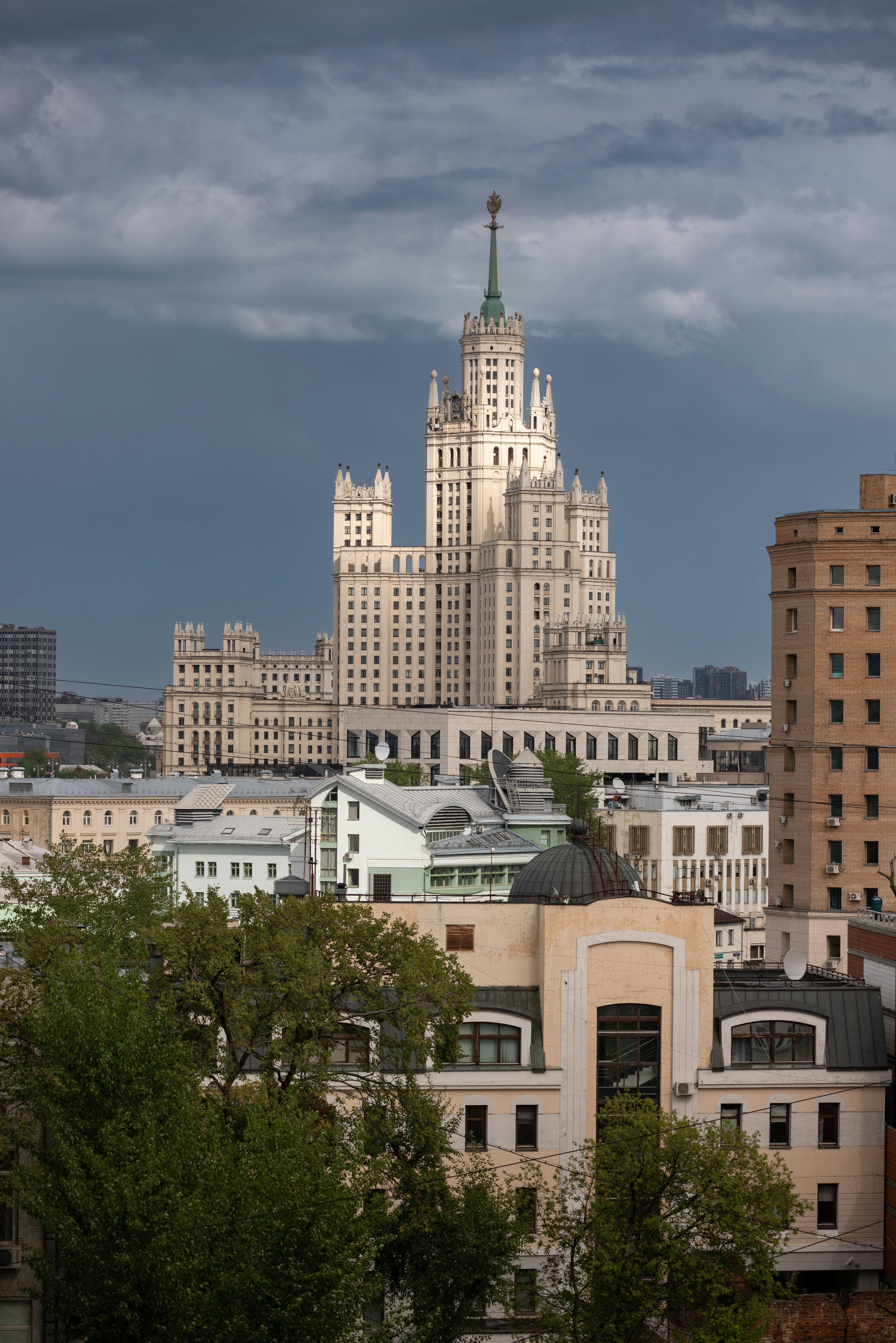 Iconic Soviet-era skyscraper towering over a cityscape under a dramatic sky. The architectural details contrast with contemporary buildings below.