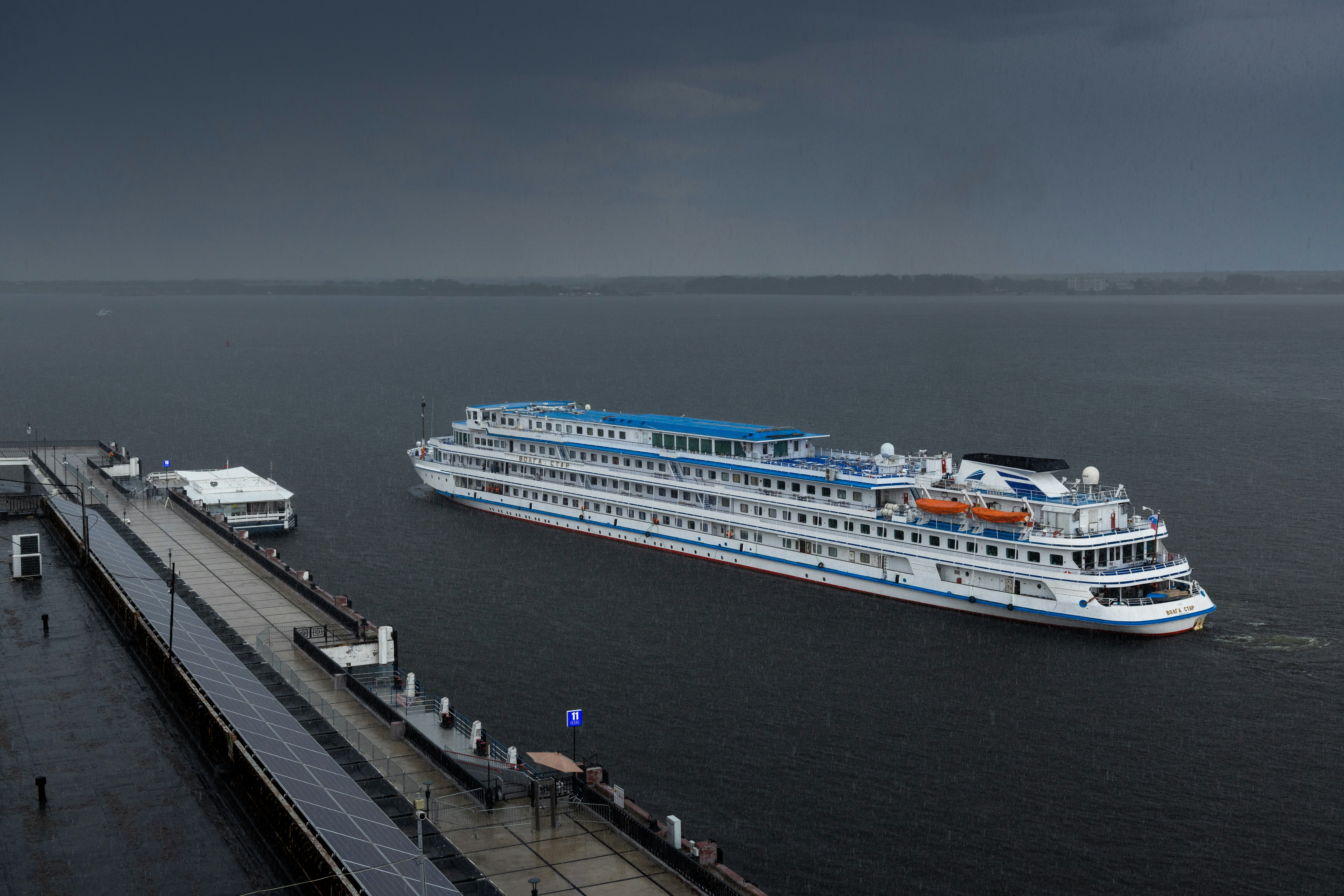 A large river cruise ship glides through dark waters under an ominous sky, with a distant pier visible. The scene captures the contrast between the vessel's bright colors and the brooding atmosphere.