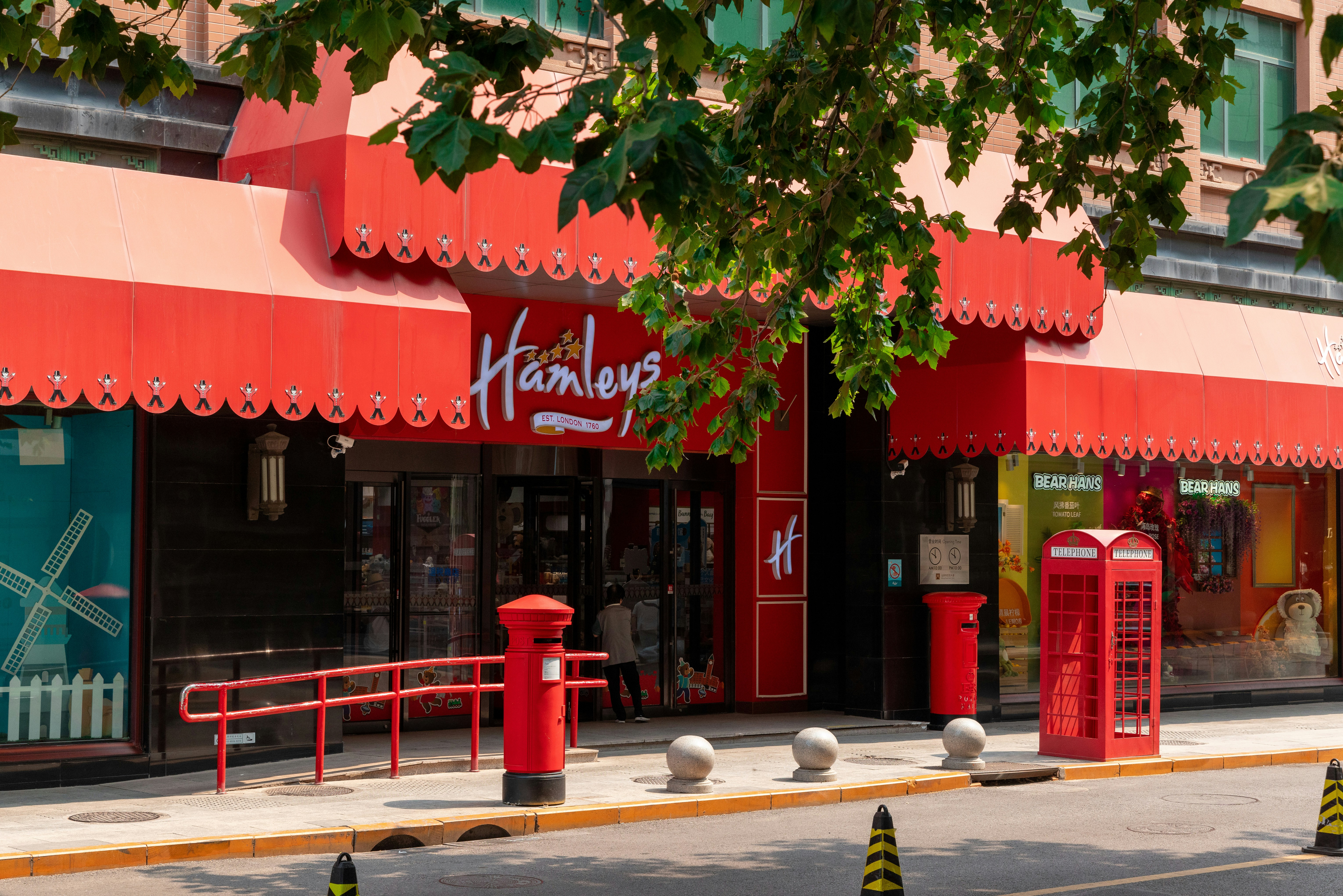 Vibrant storefront of Hamleys toy store featuring red awnings and iconic red telephone boxes, framed by lush green foliage.