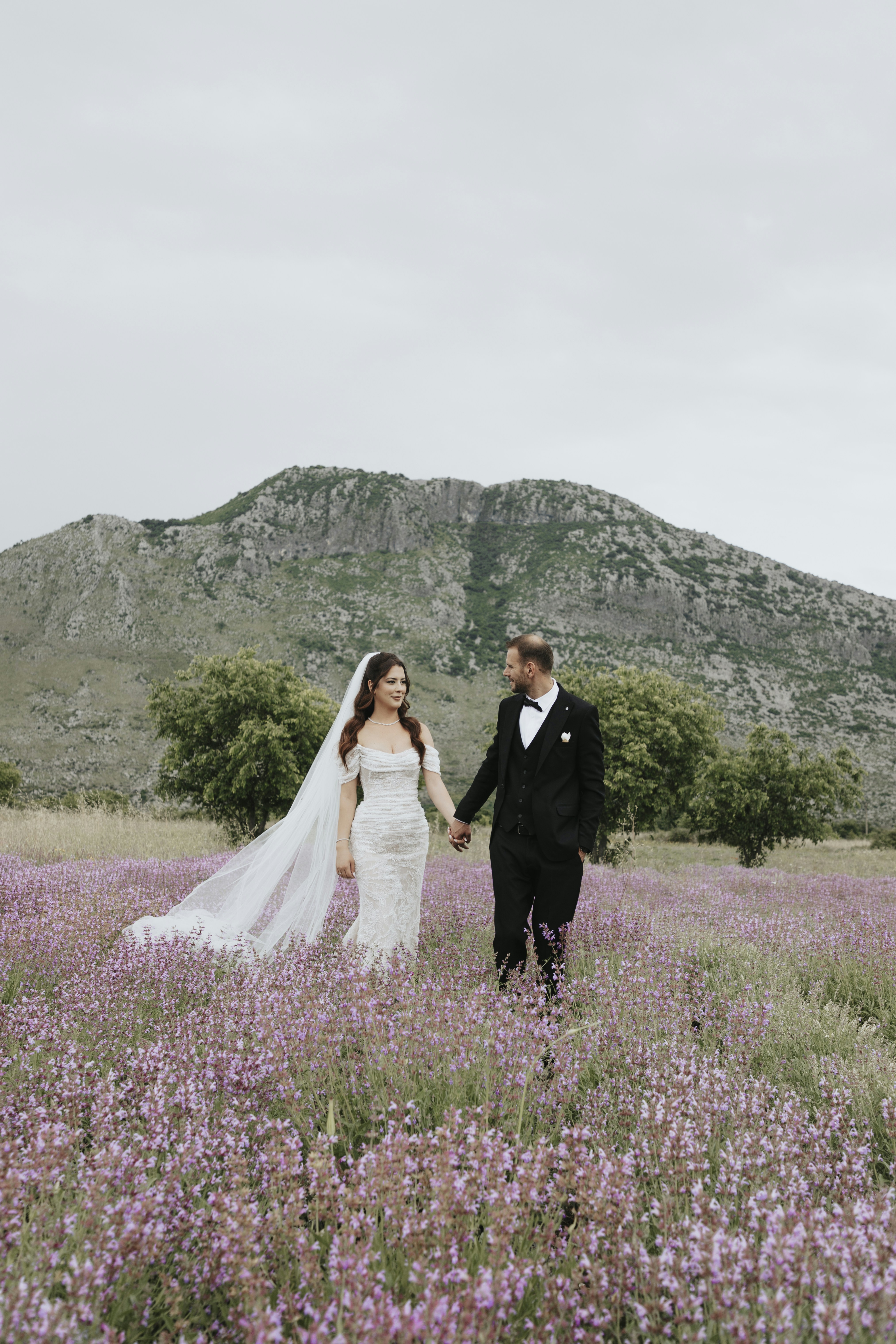 Couple walking hand in hand through a vibrant field of lavender flowers with a mountainous backdrop.