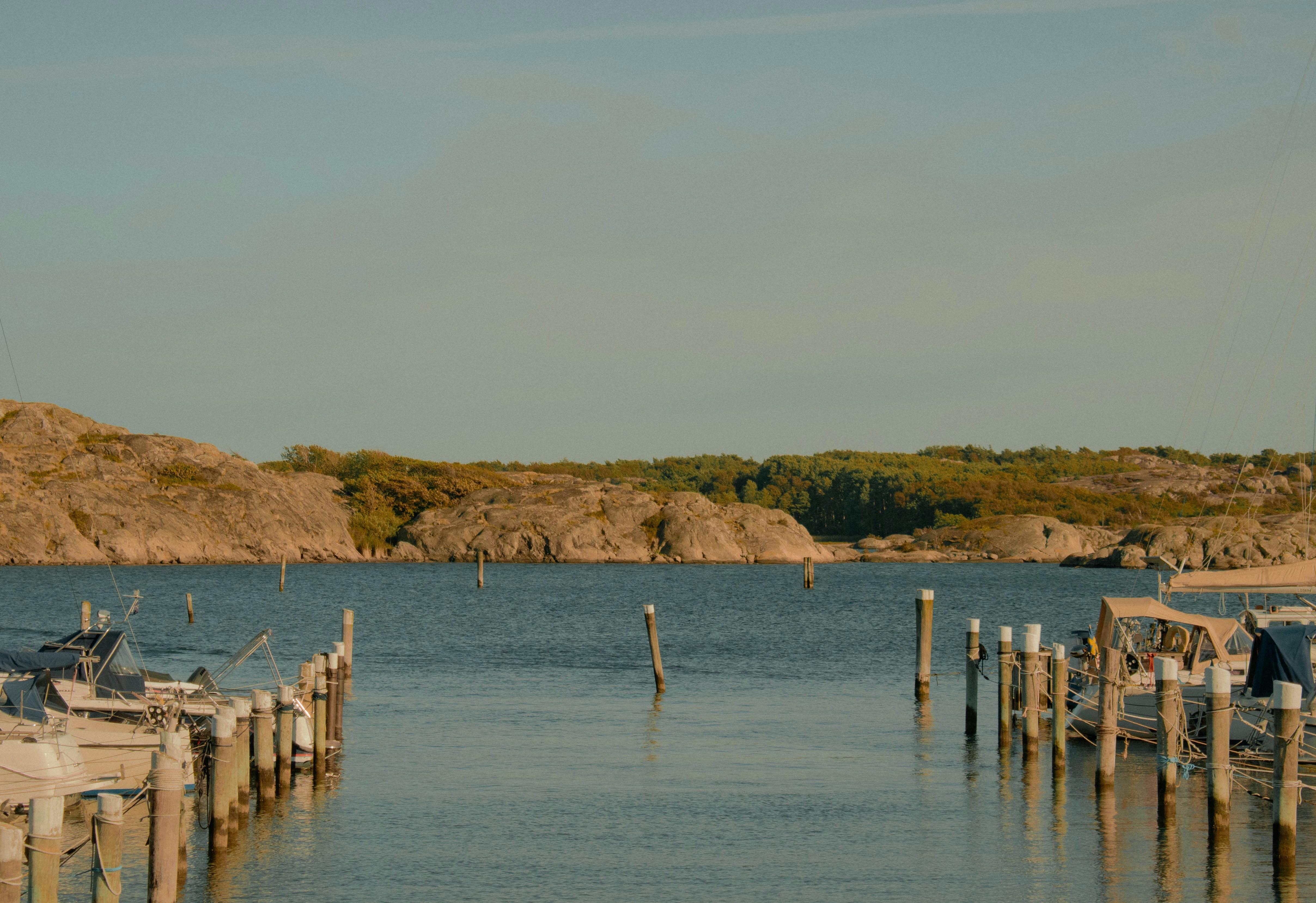 Boats moored along a tranquil harbor, framed by rugged coastal rocks and lush greenery. The calm water reflects the soft hues of the sky.