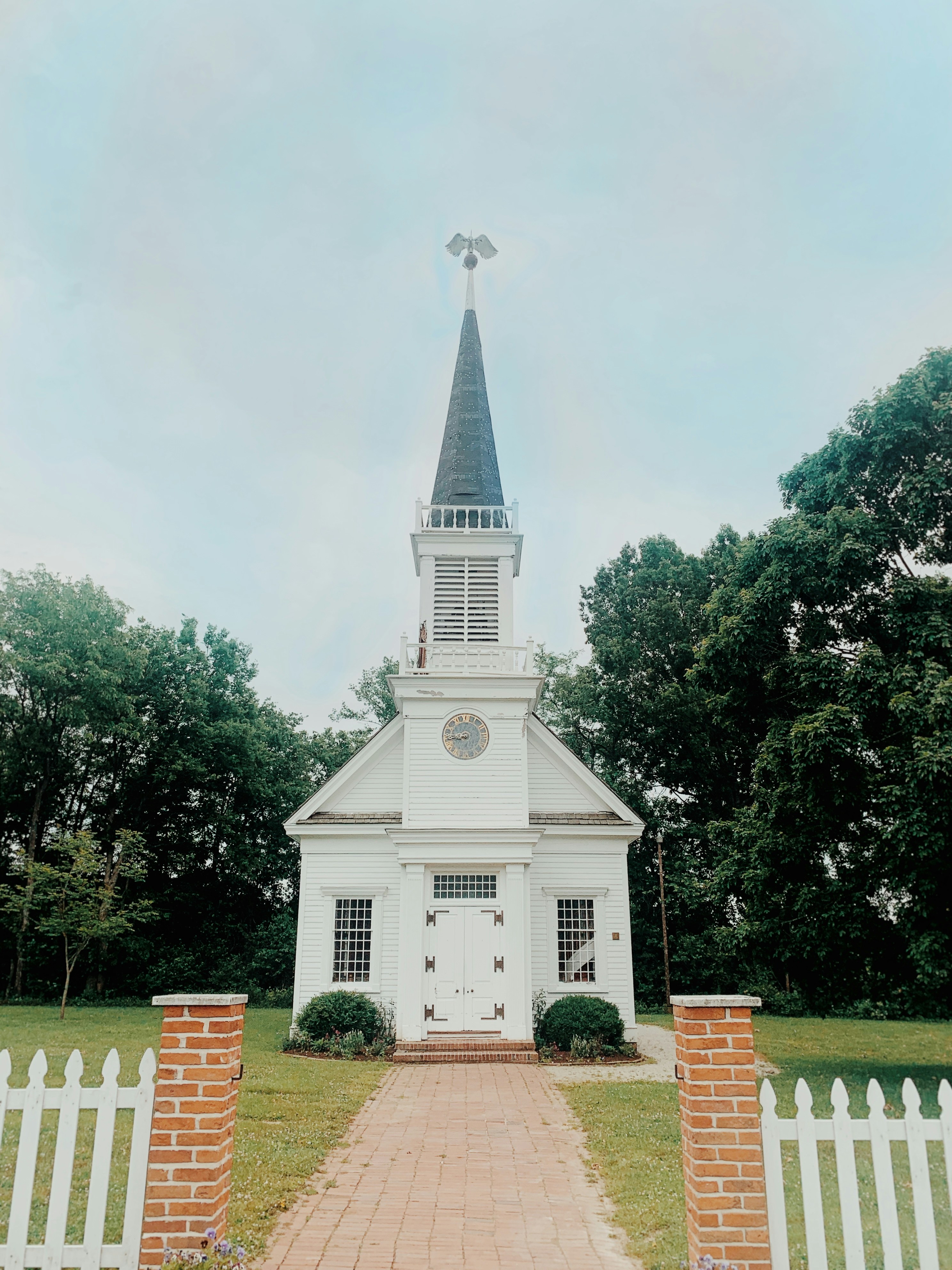 Charming white chapel with a tall steeple, framed by lush greenery and a quaint picket fence. The serene setting invites reflection.