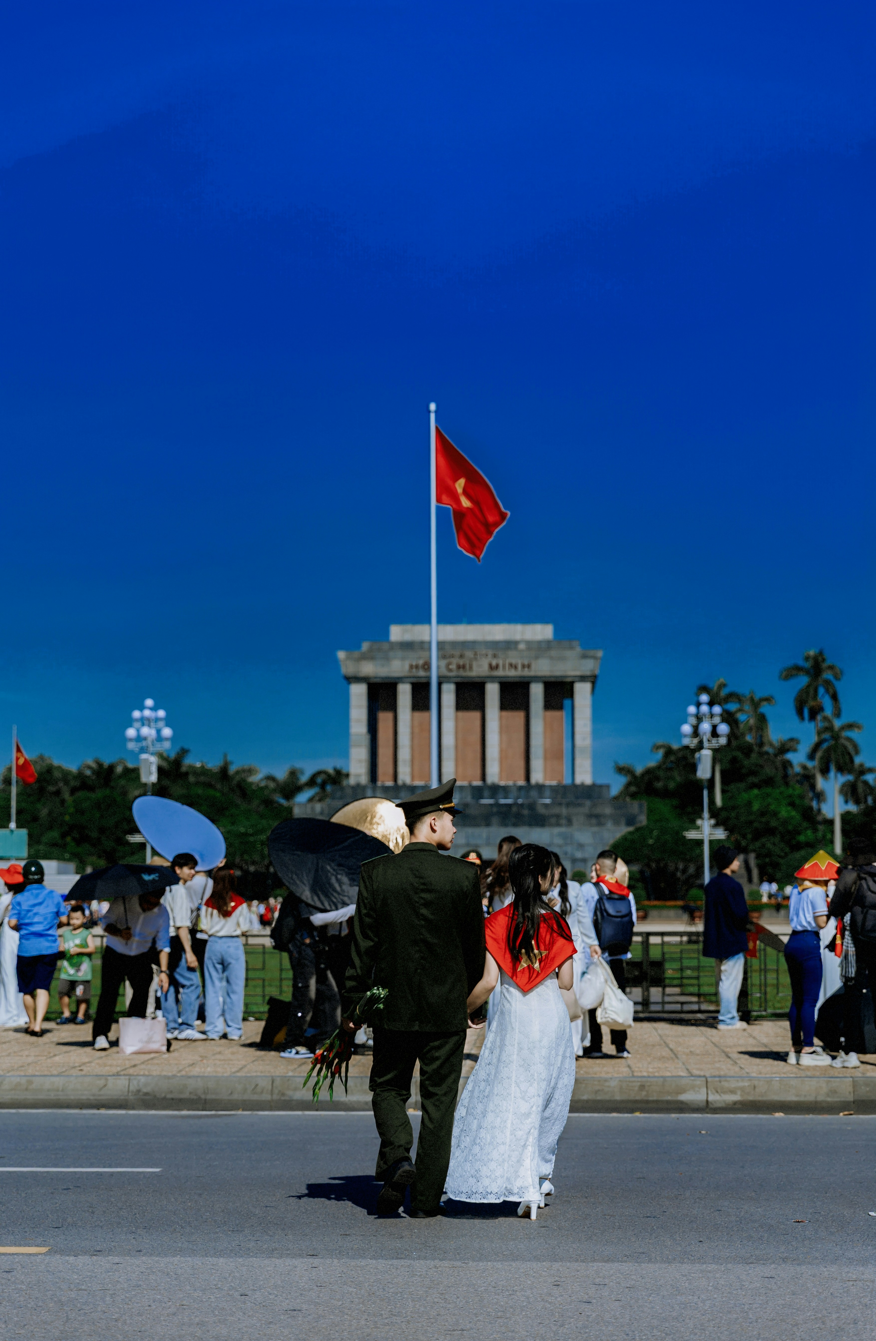 A couple in traditional attire walks hand-in-hand, with Ho Chi Minh's mausoleum and the Vietnamese flag in the background, amidst a gathering of onlookers.