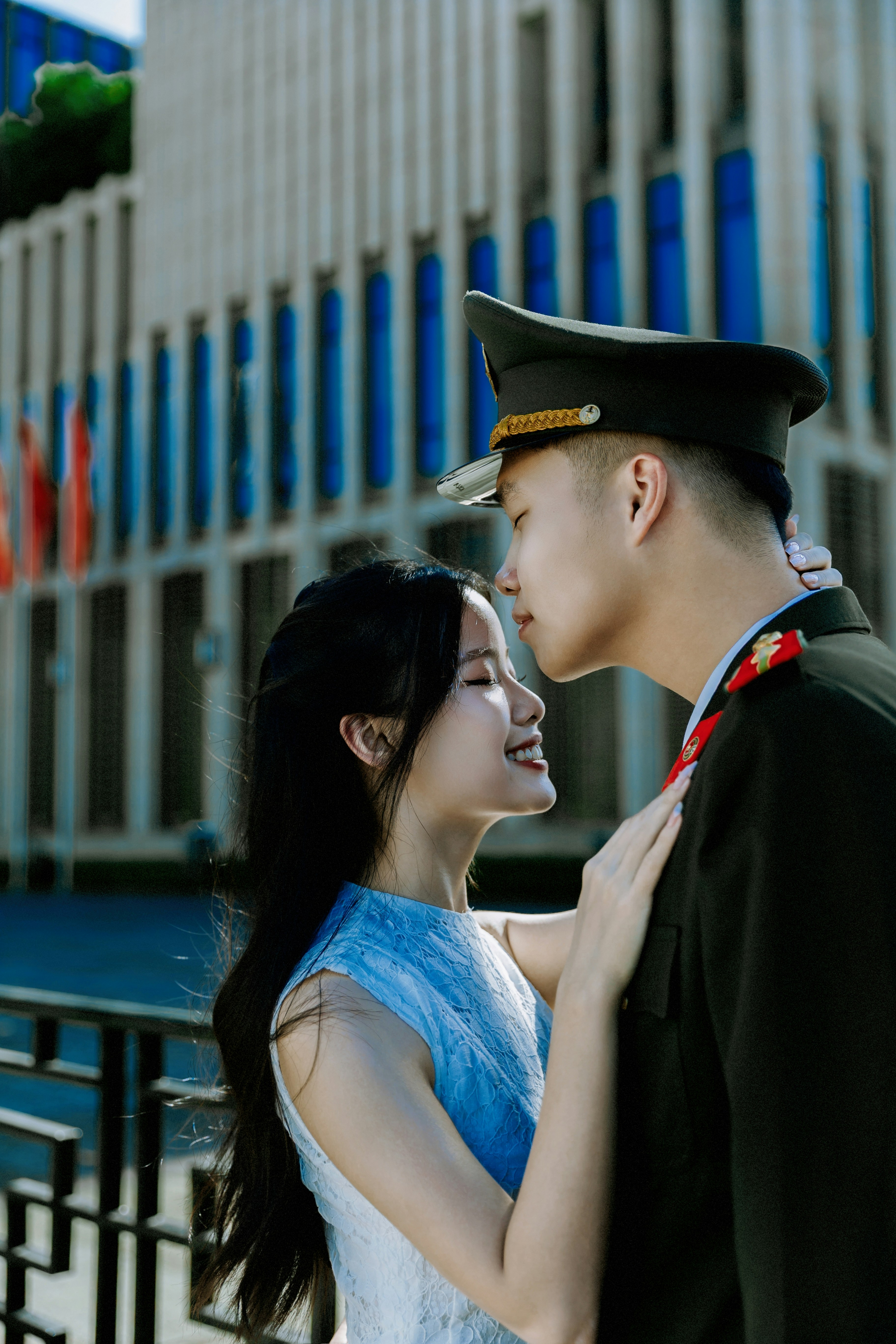 Couple sharing a tender moment, with the woman in a light blue dress and the man in military uniform, set against a backdrop of flags and modern architecture.