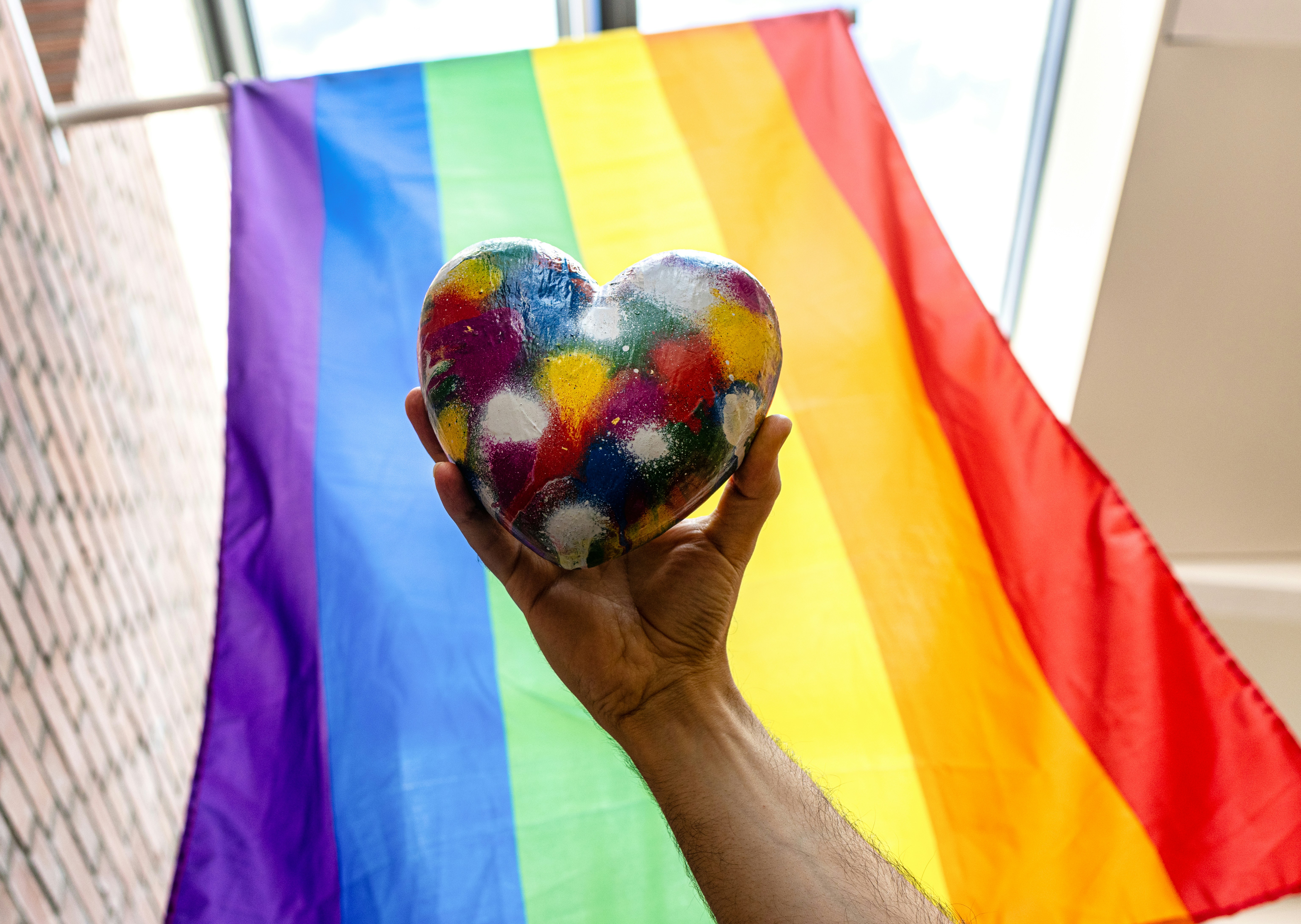Colorful heart-shaped object held against a backdrop of a rainbow flag, symbolizing pride and inclusivity.