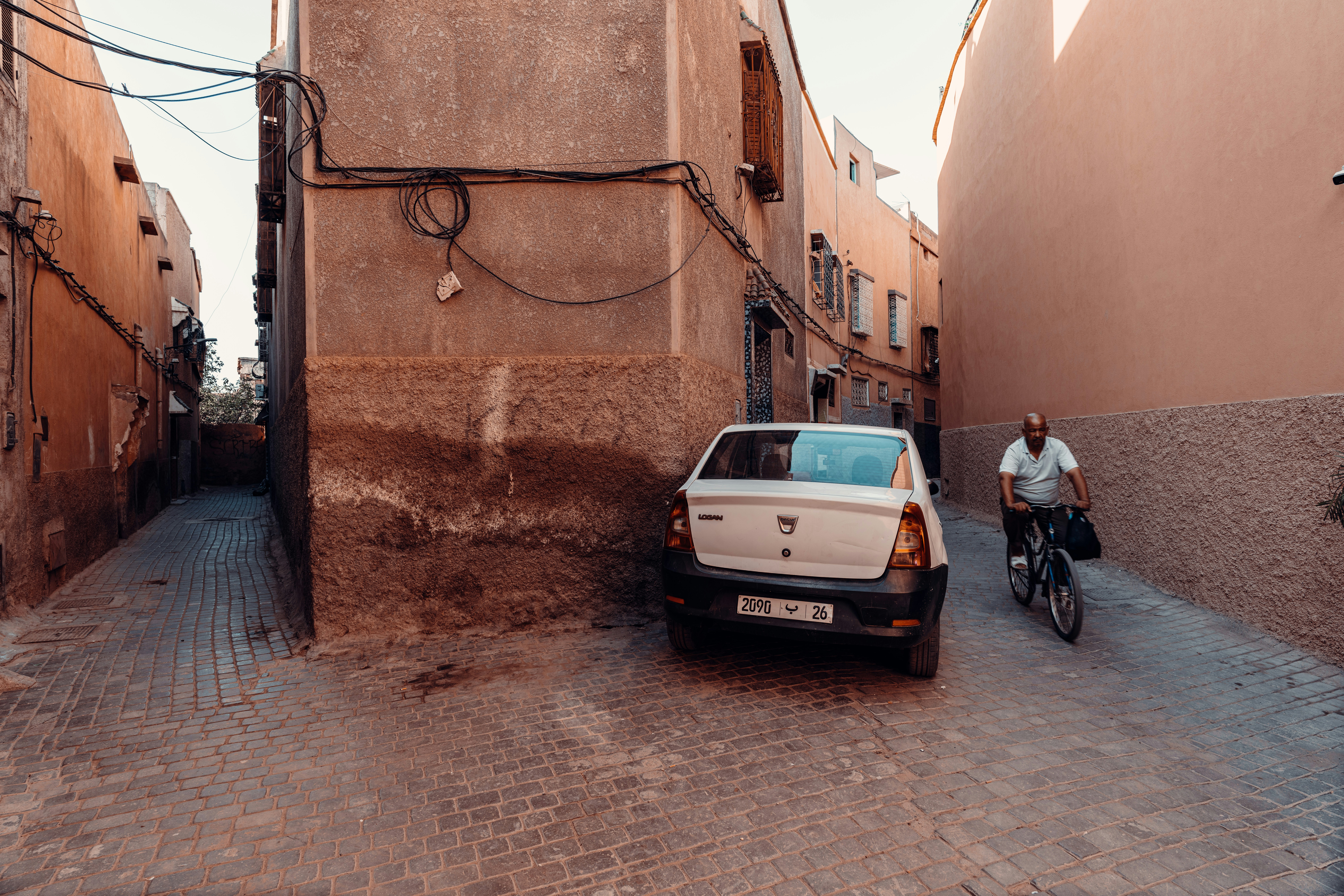 Man rides bicycle down narrow street with car.