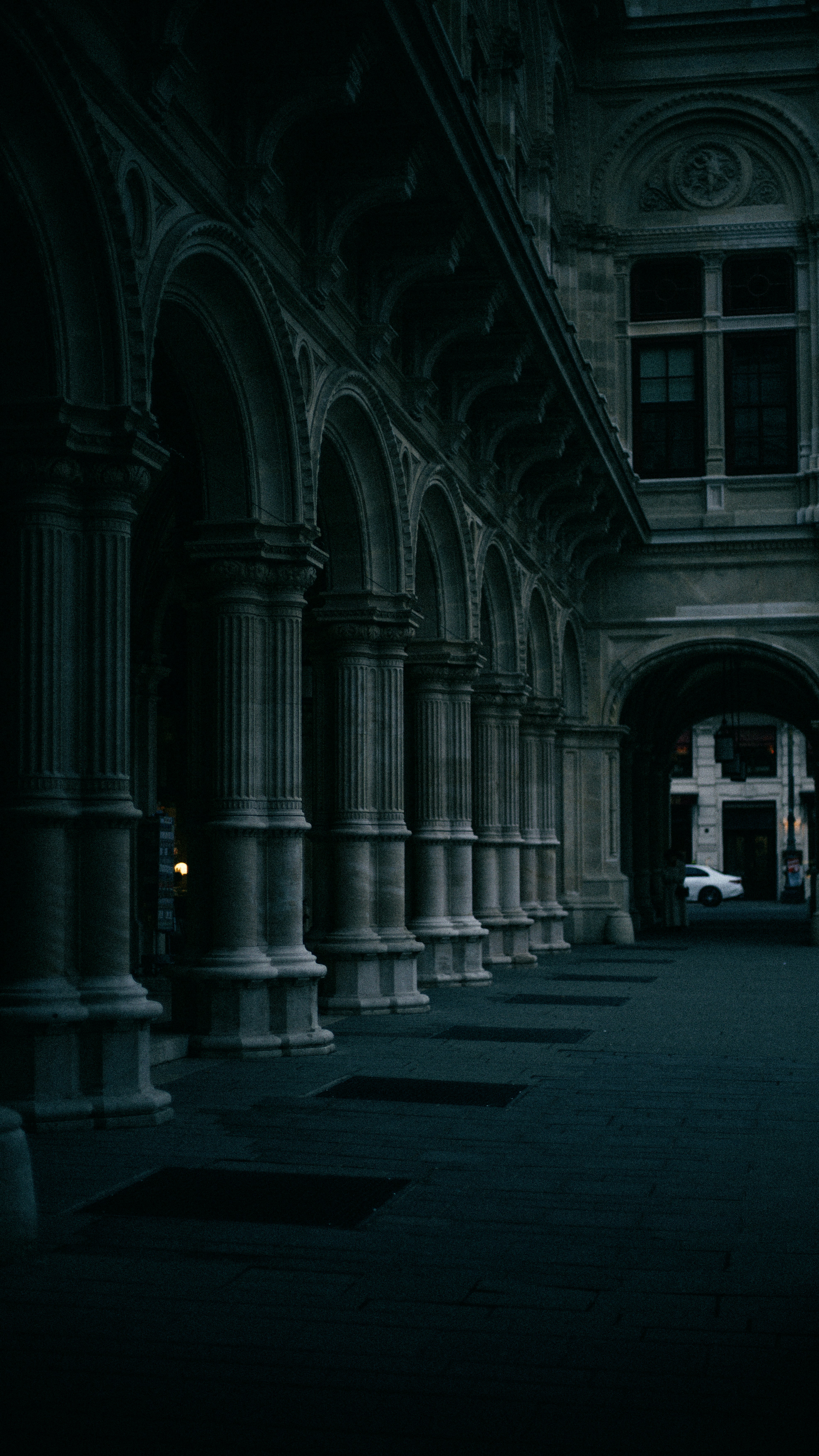A moody evening view of the Vienna State Opera’s historic arcade with classical columns and arches. The dim light and vintage tones create a cinematic atmosphere, highlighting the elegant European architecture and timeless charm of the city. | Ornate stone arches and columns line a dark walkway.