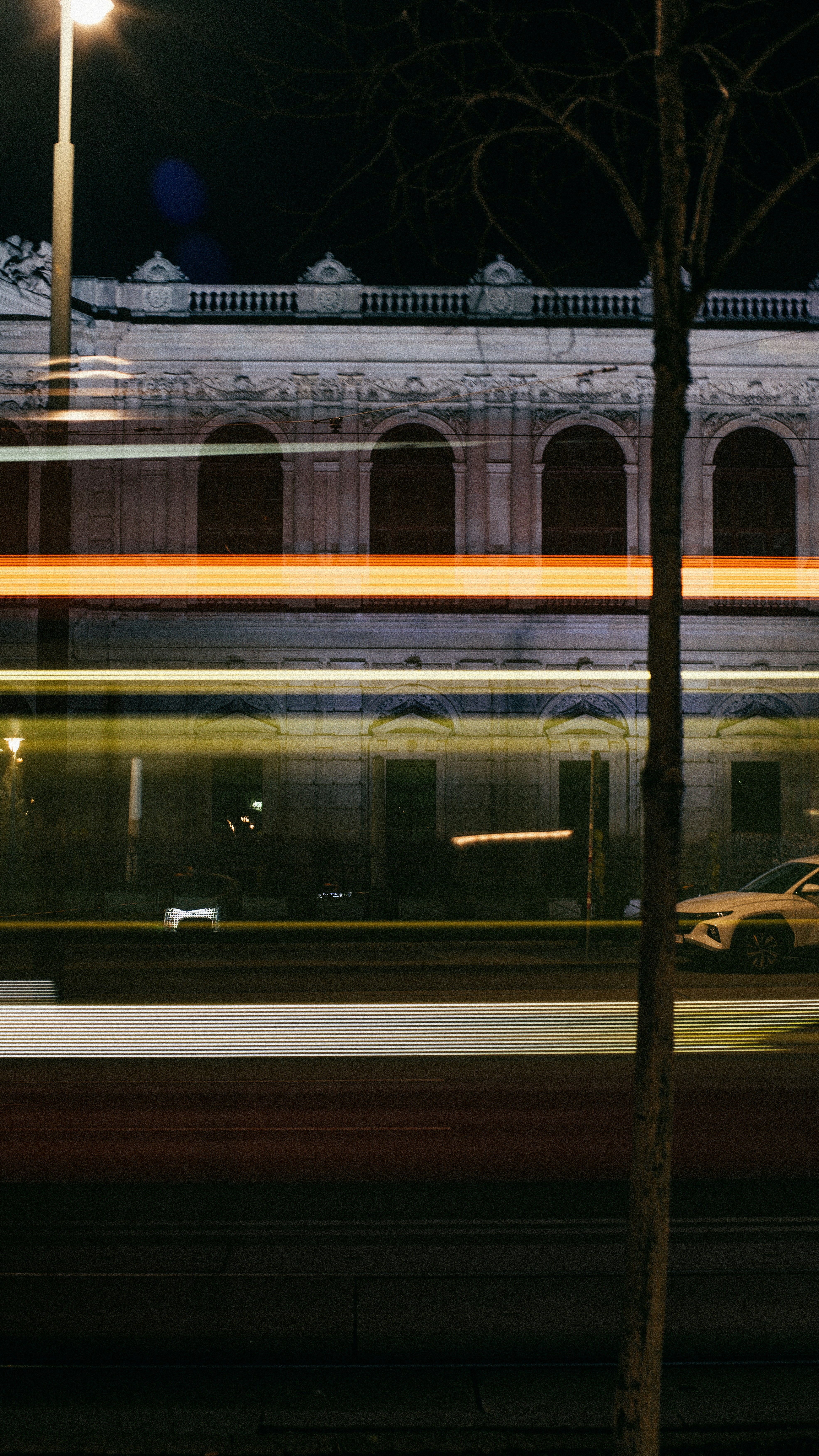 Long exposure night shot of a tram passing in front of a historic building in Vienna. The colorful light trails contrast with the classical architecture, capturing the essence of modern urban life meeting European heritage in a cinematic atmosphere. | City street at night with light trails and car trails.
