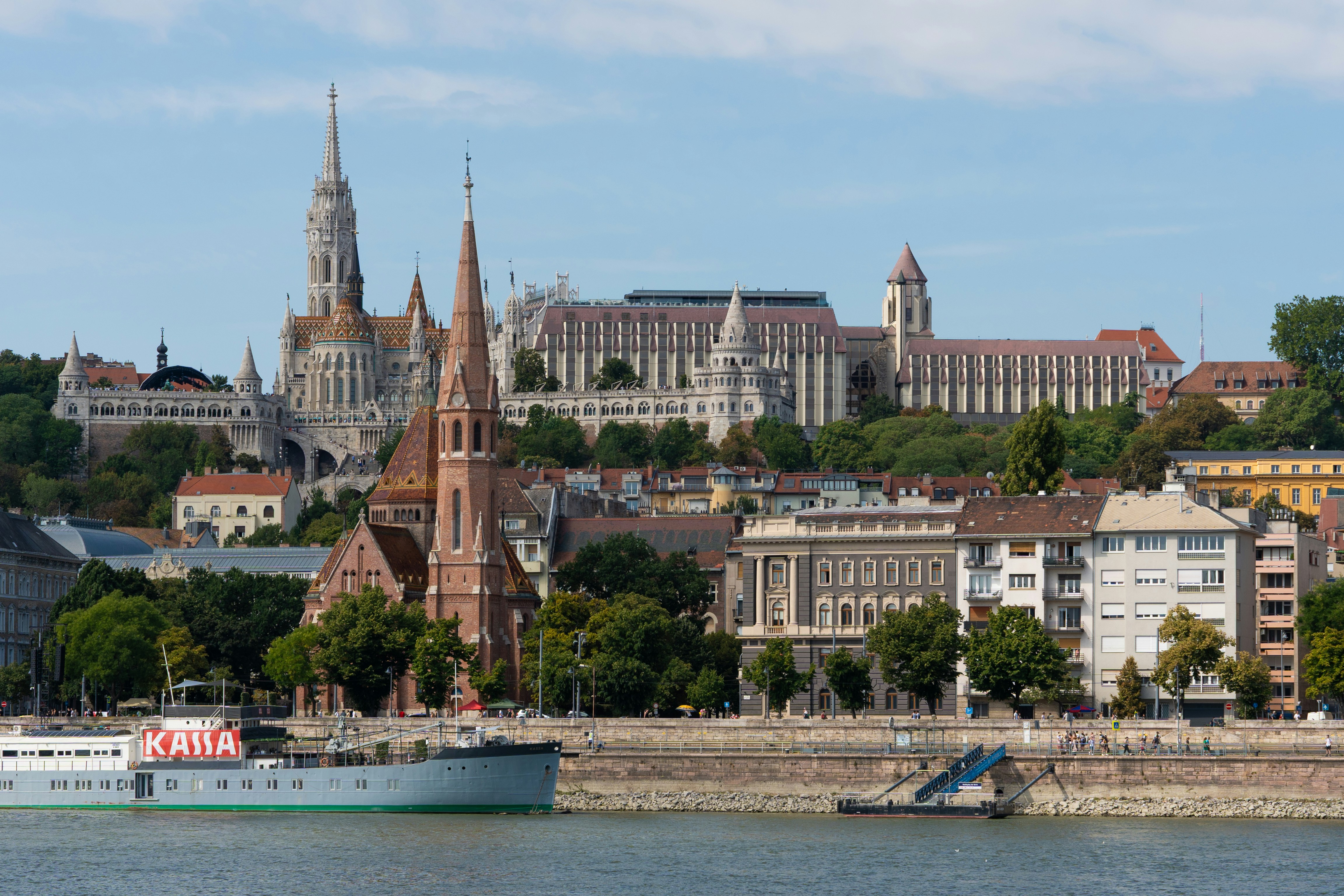 Historic buildings and a church line the banks of the Danube River, with a boat in the foreground. The scene captures a blend of architectural styles against a vibrant backdrop.