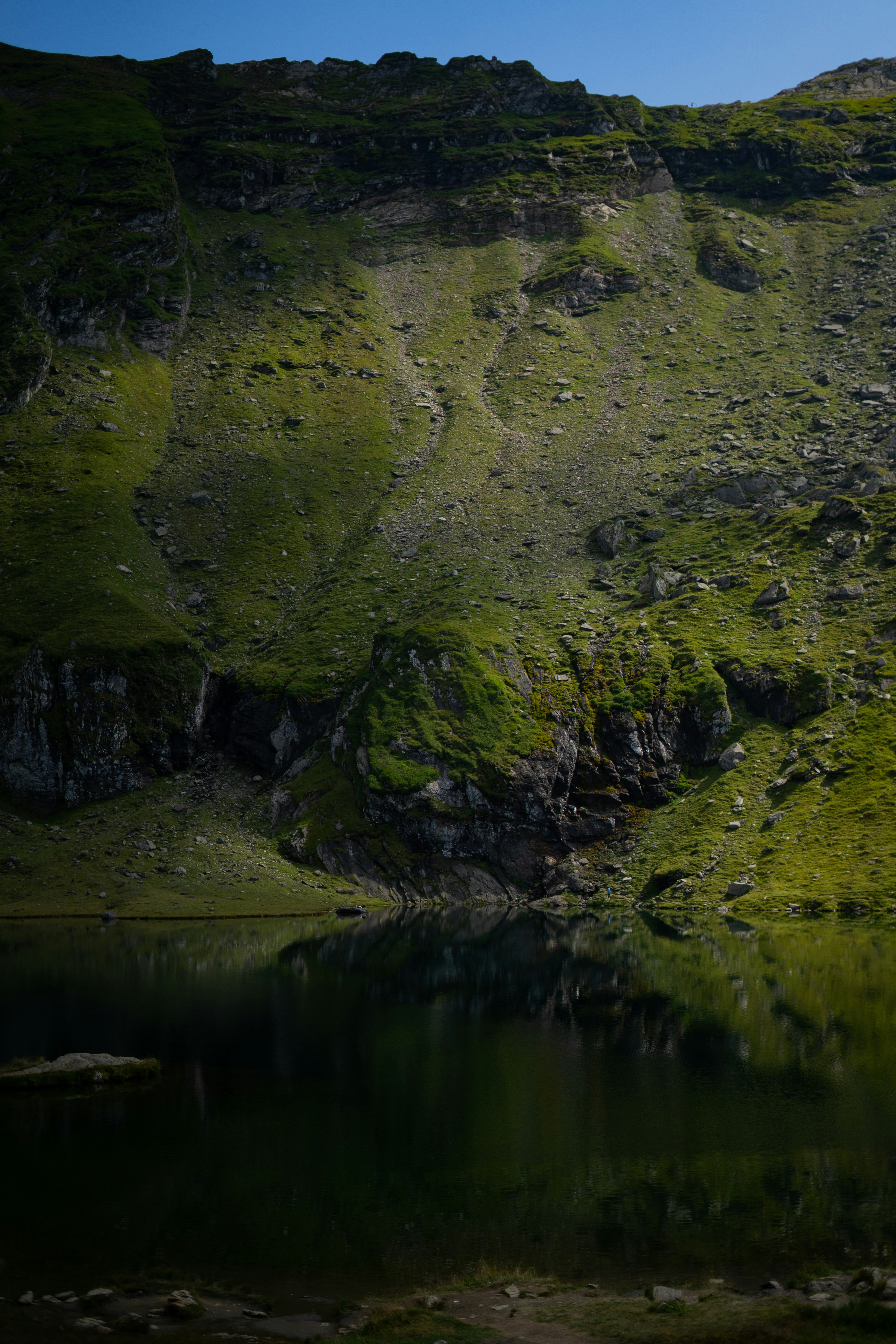Calm lake reflects a rugged, green mountain slope.