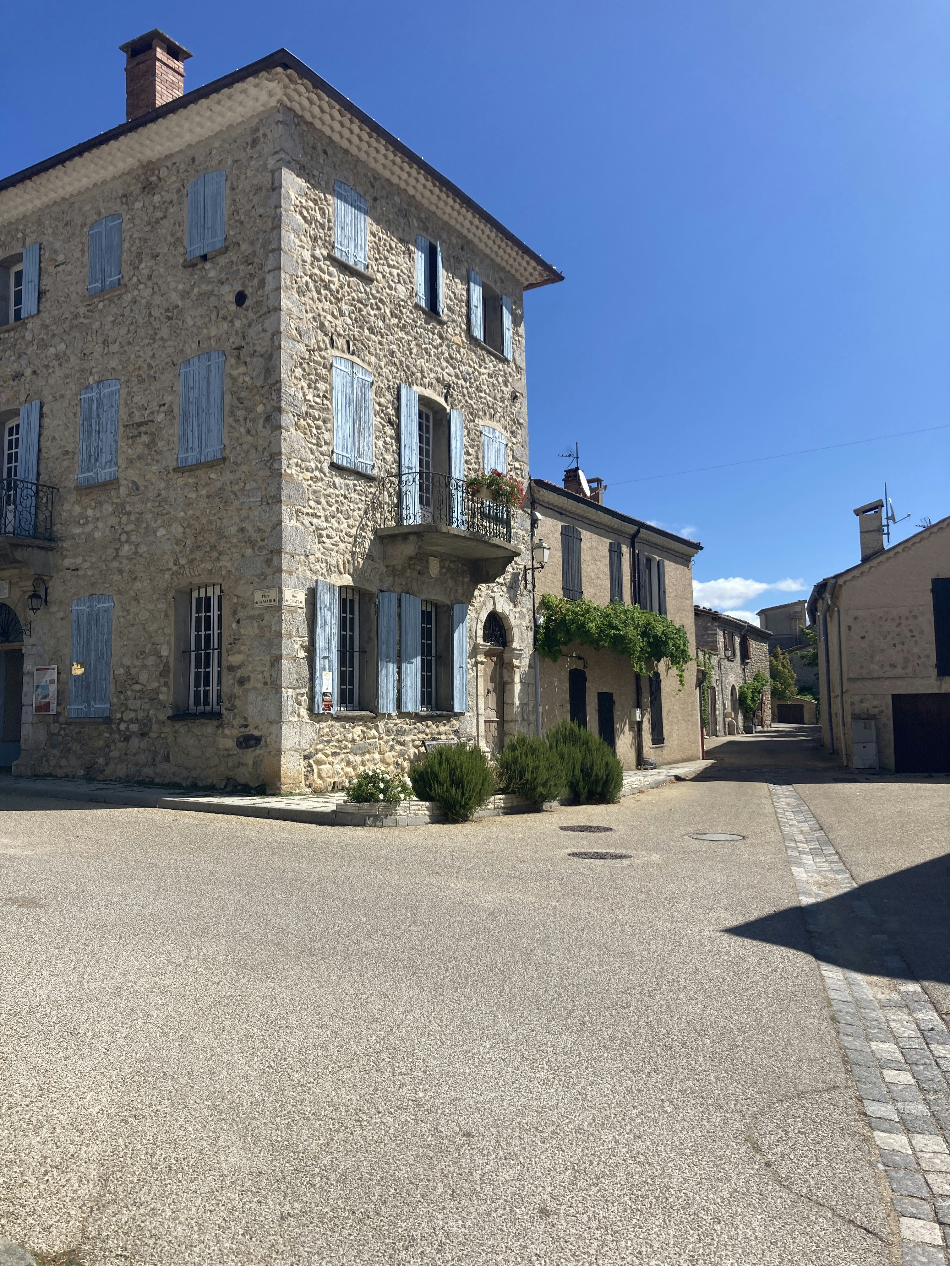 Historic stone building with blue shutters and a balcony, surrounded by a picturesque village street.
