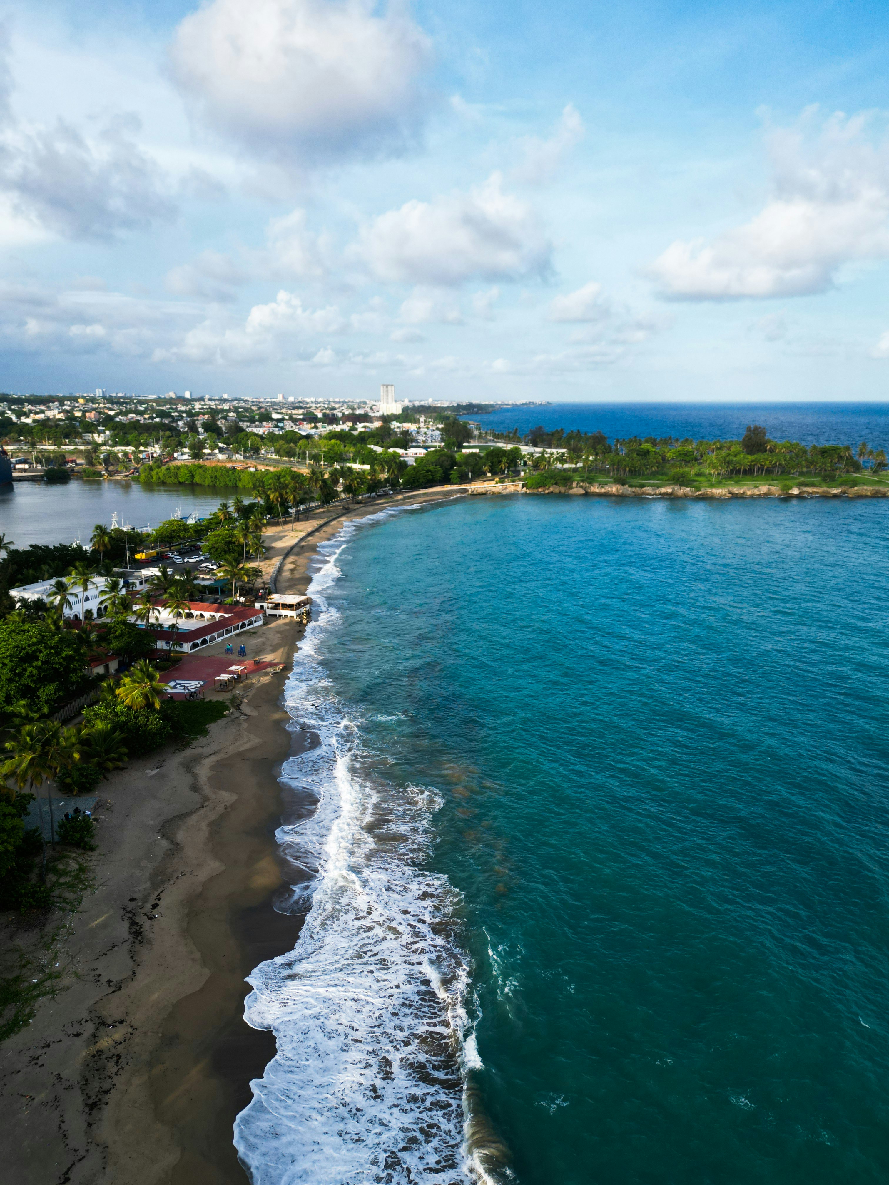 Aerial view capturing the coastline where lush greenery meets vibrant blue waters, showcasing a tranquil beach and nearby urban landscape.