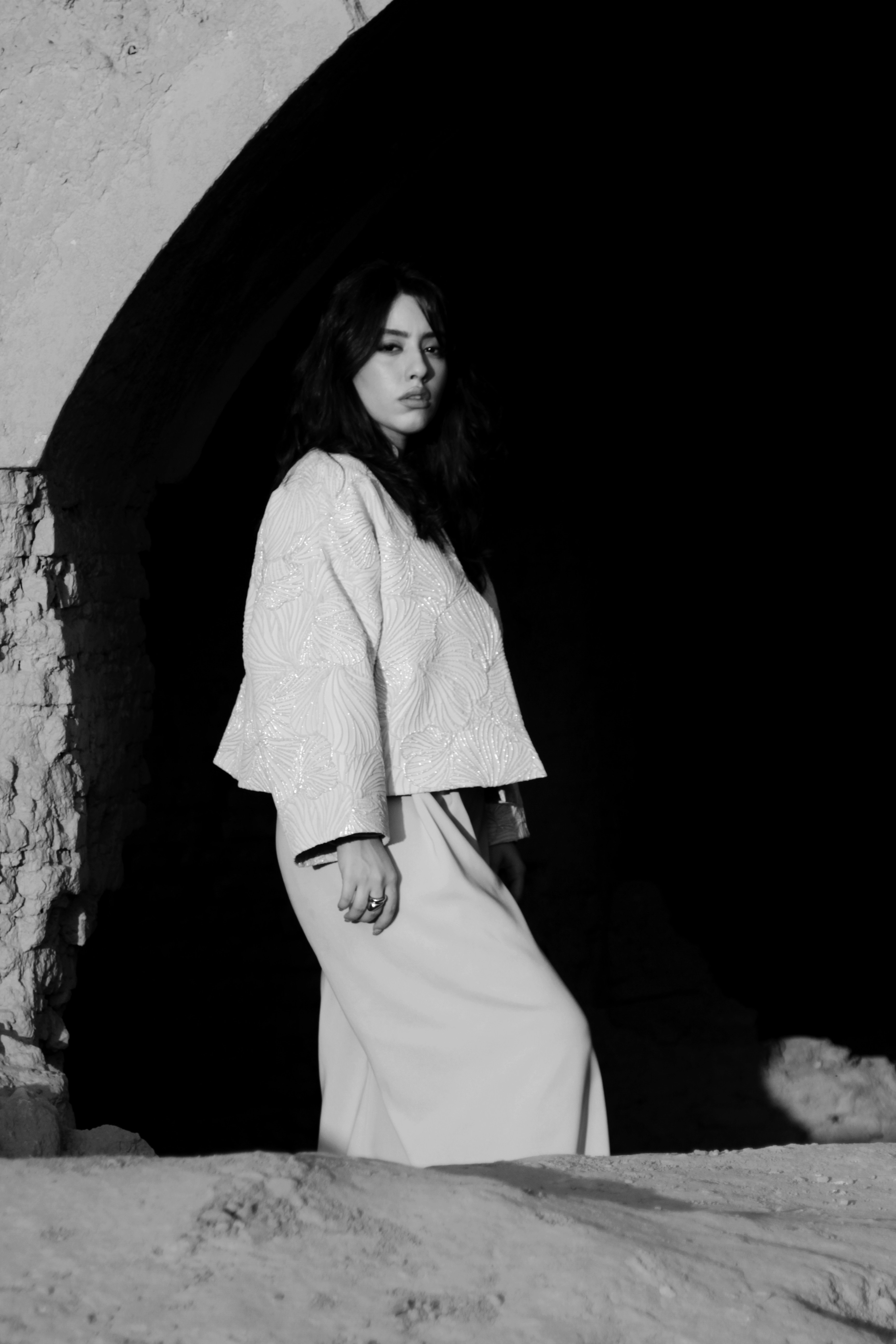 A woman stands elegantly in a dimly lit archway, her expression contemplative against a stark contrast of light and shadow.