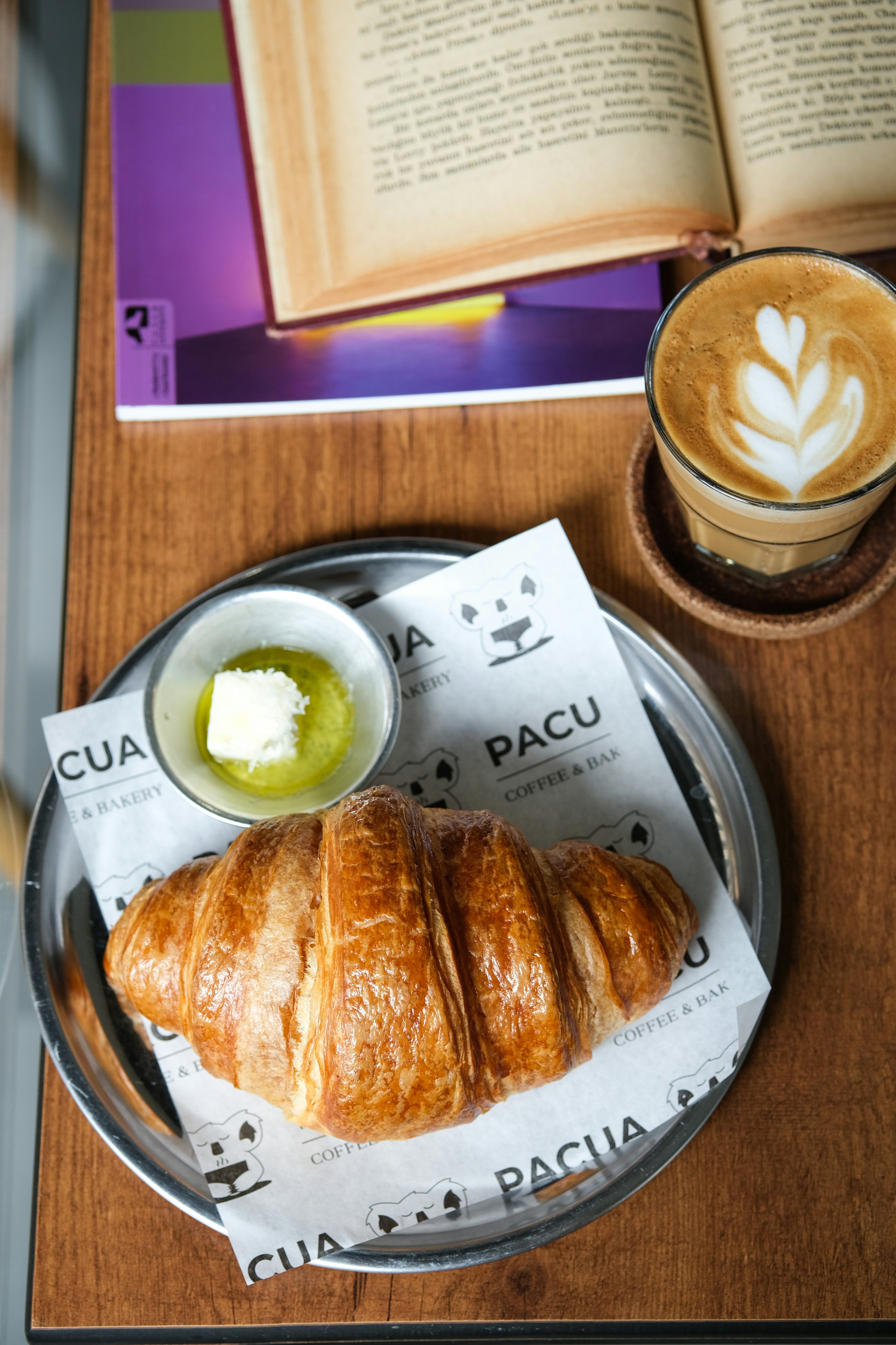 Golden croissant accompanied by a small bowl of butter and a beautifully crafted latte on a wooden table, with an open book in the background.