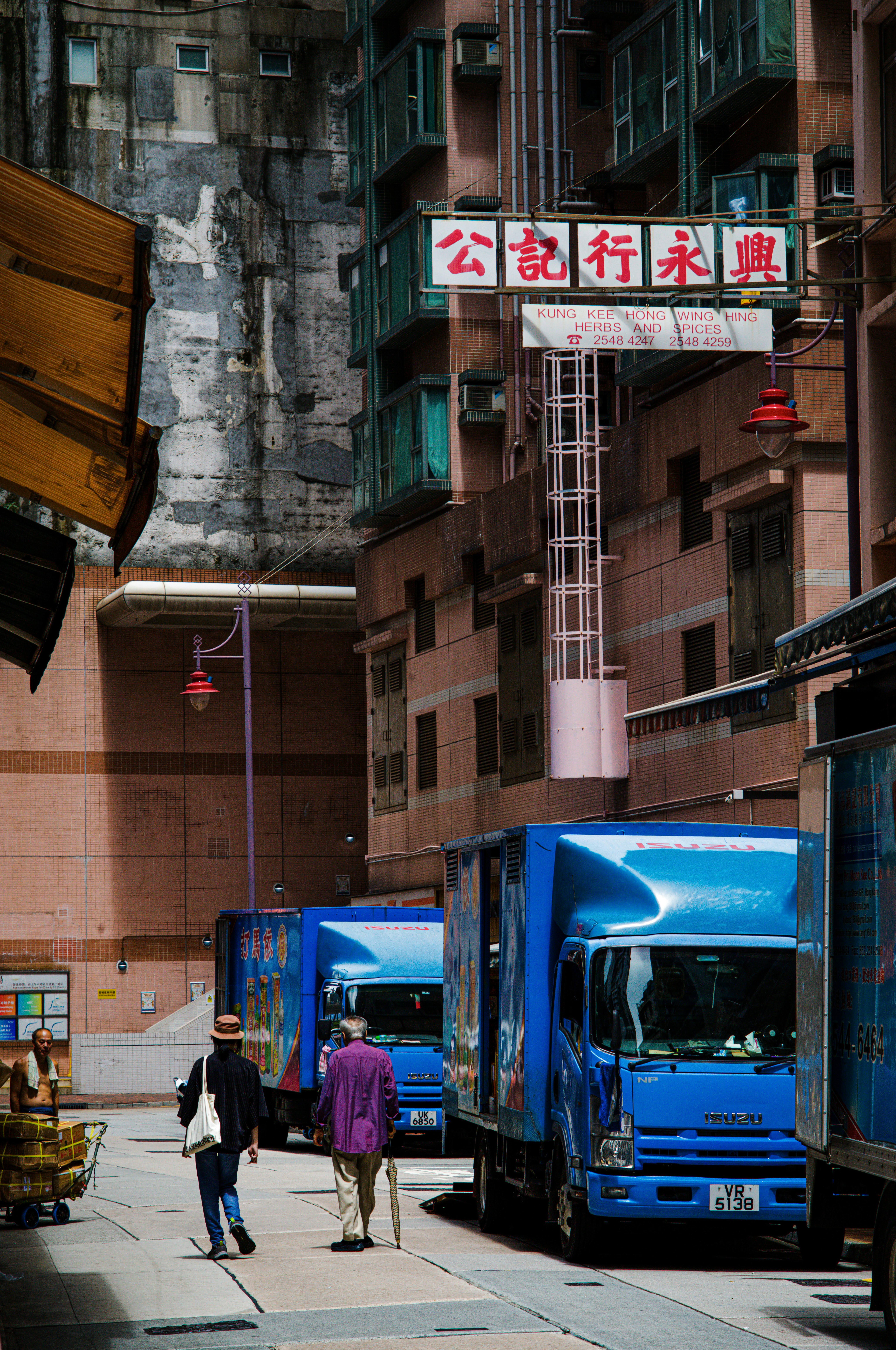 Two people walk past blue trucks on a street.