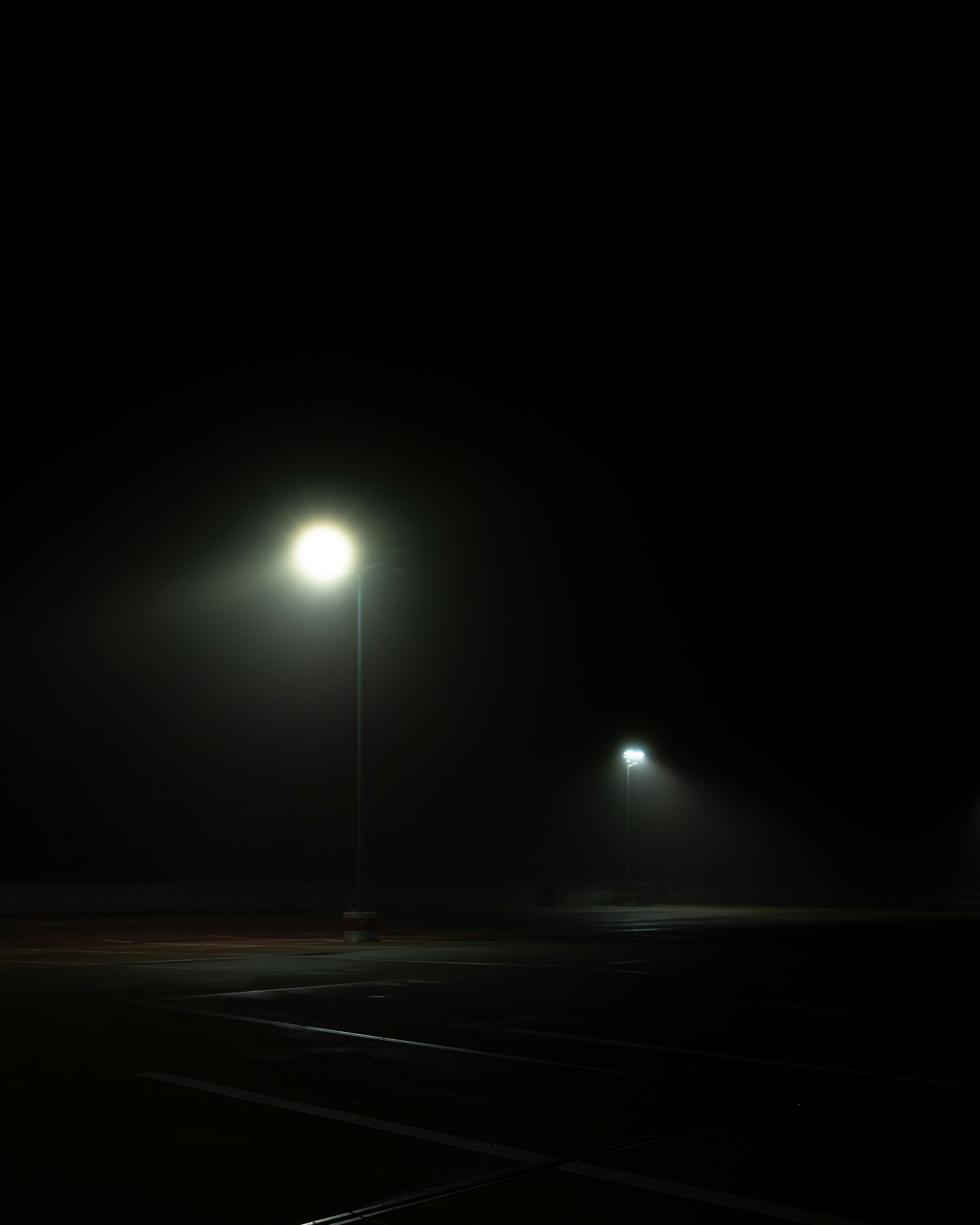 Two streetlights casting a soft glow in an empty, foggy parking lot at night.
