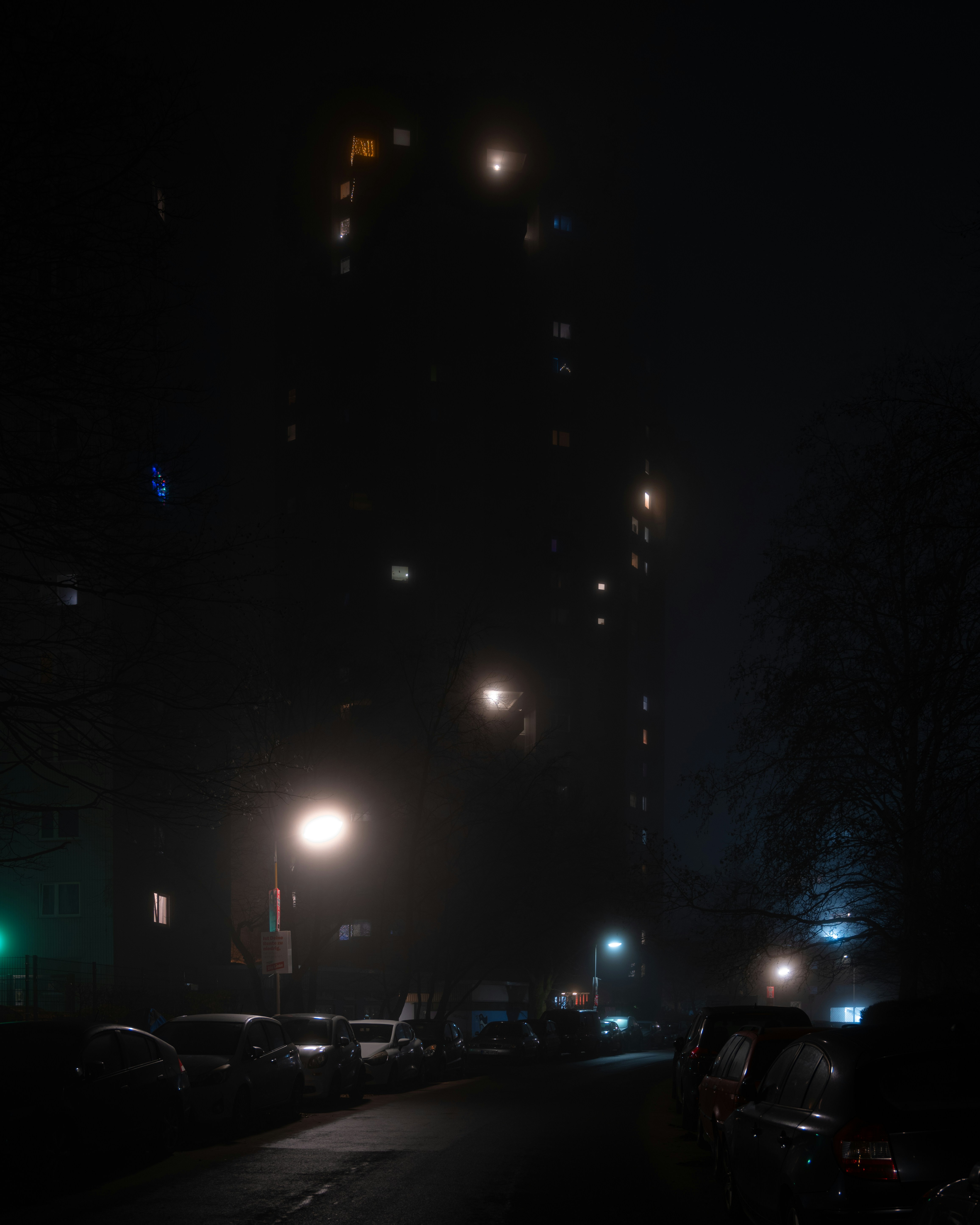 Dimly lit urban street shrouded in fog, with glowing windows in a distant high-rise and soft streetlights illuminating parked cars.