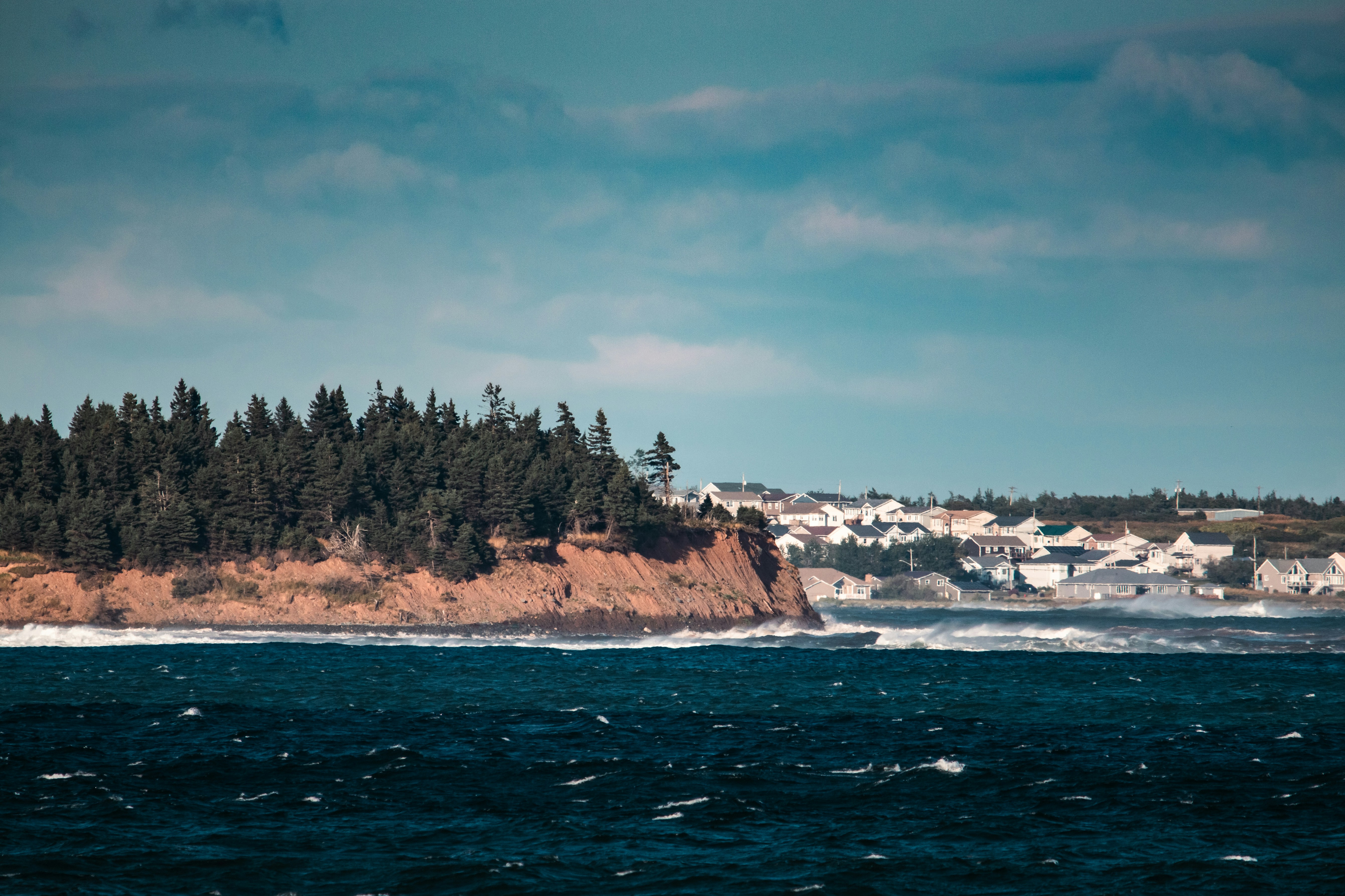 Lush green cliffs rise above churning waves, leading to a quaint coastal village nestled in the distance. The scene captures the serene interplay between nature and human habitation.