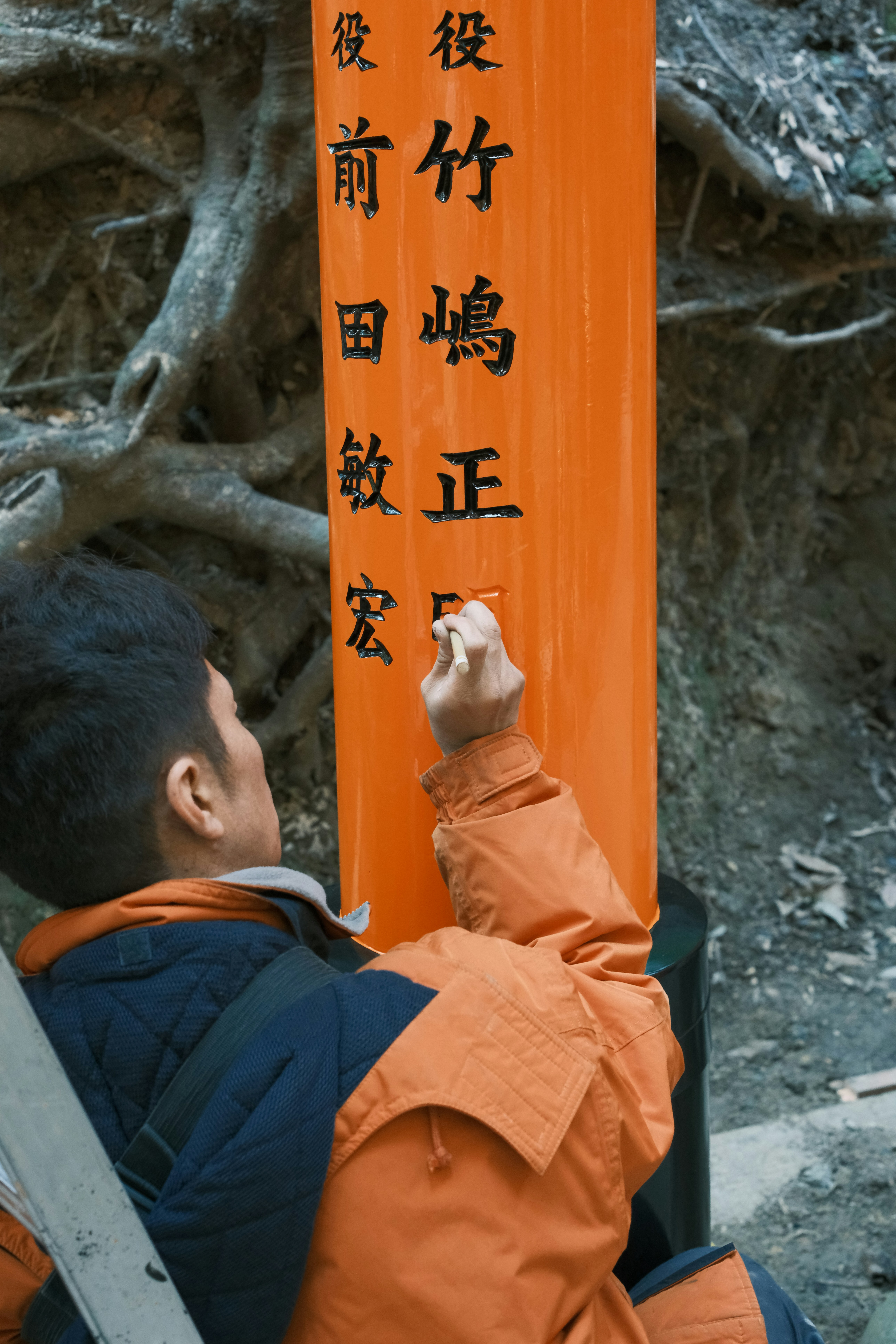 A person in an orange jacket meticulously writes on a vibrant orange post, showcasing traditional calligraphy techniques in a natural setting.