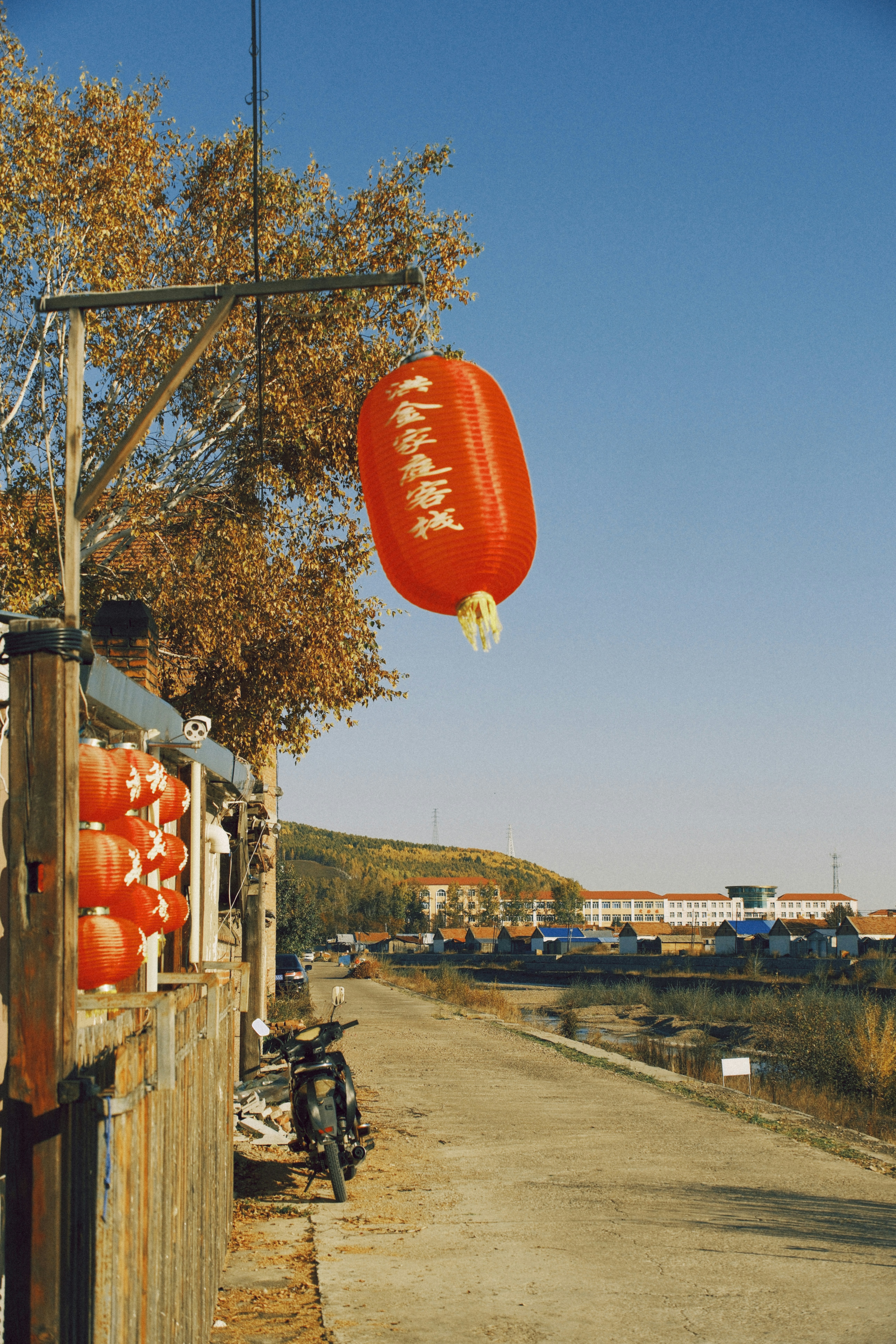 Vibrant red lanterns adorn a rustic wooden structure beside a tranquil dirt path leading to a serene river, framed by autumn foliage. The scene captures a harmonious blend of culture and nature.