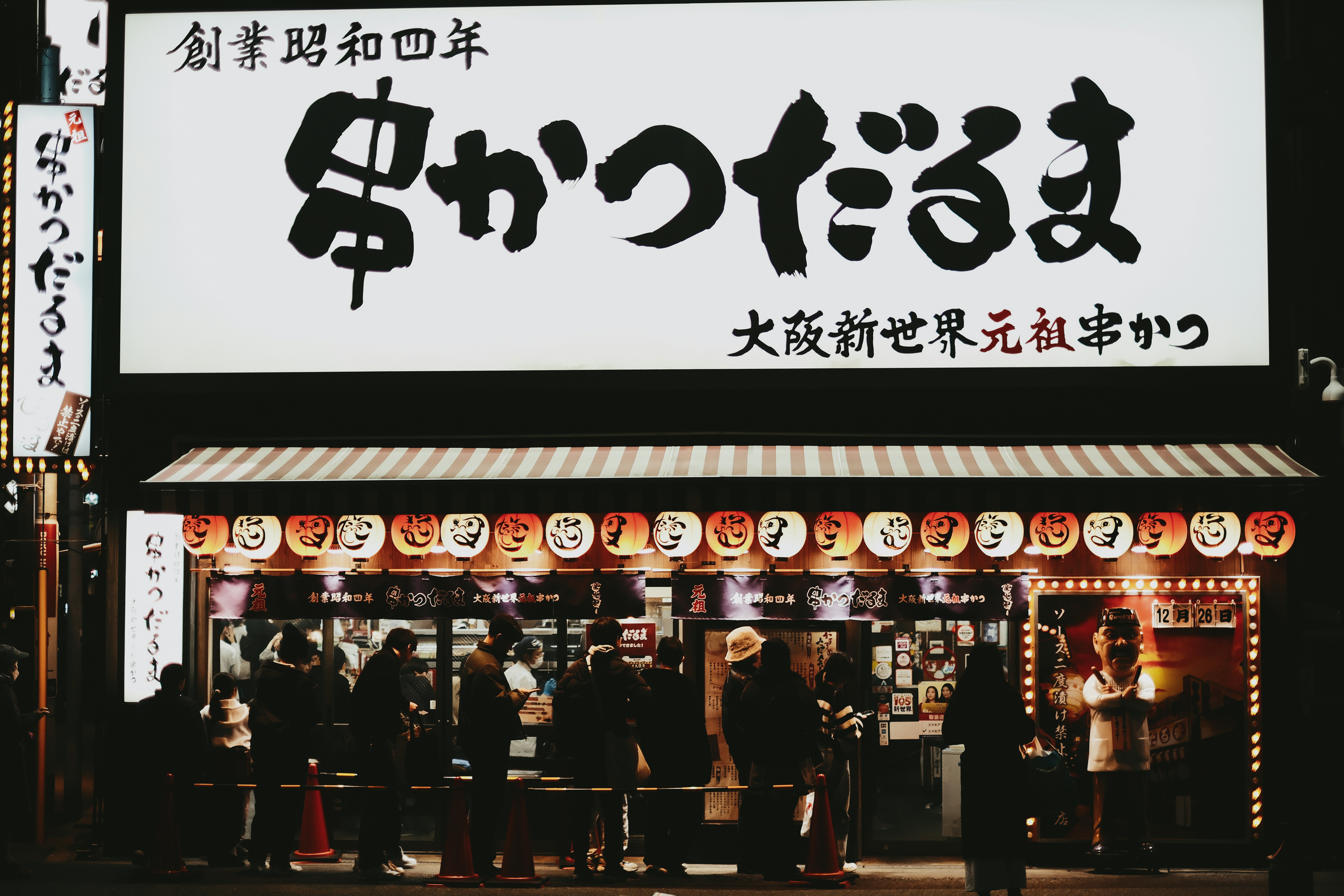 Busy street food stall in Osaka featuring vibrant lanterns and a bold sign in Japanese. Diners eagerly await their orders.