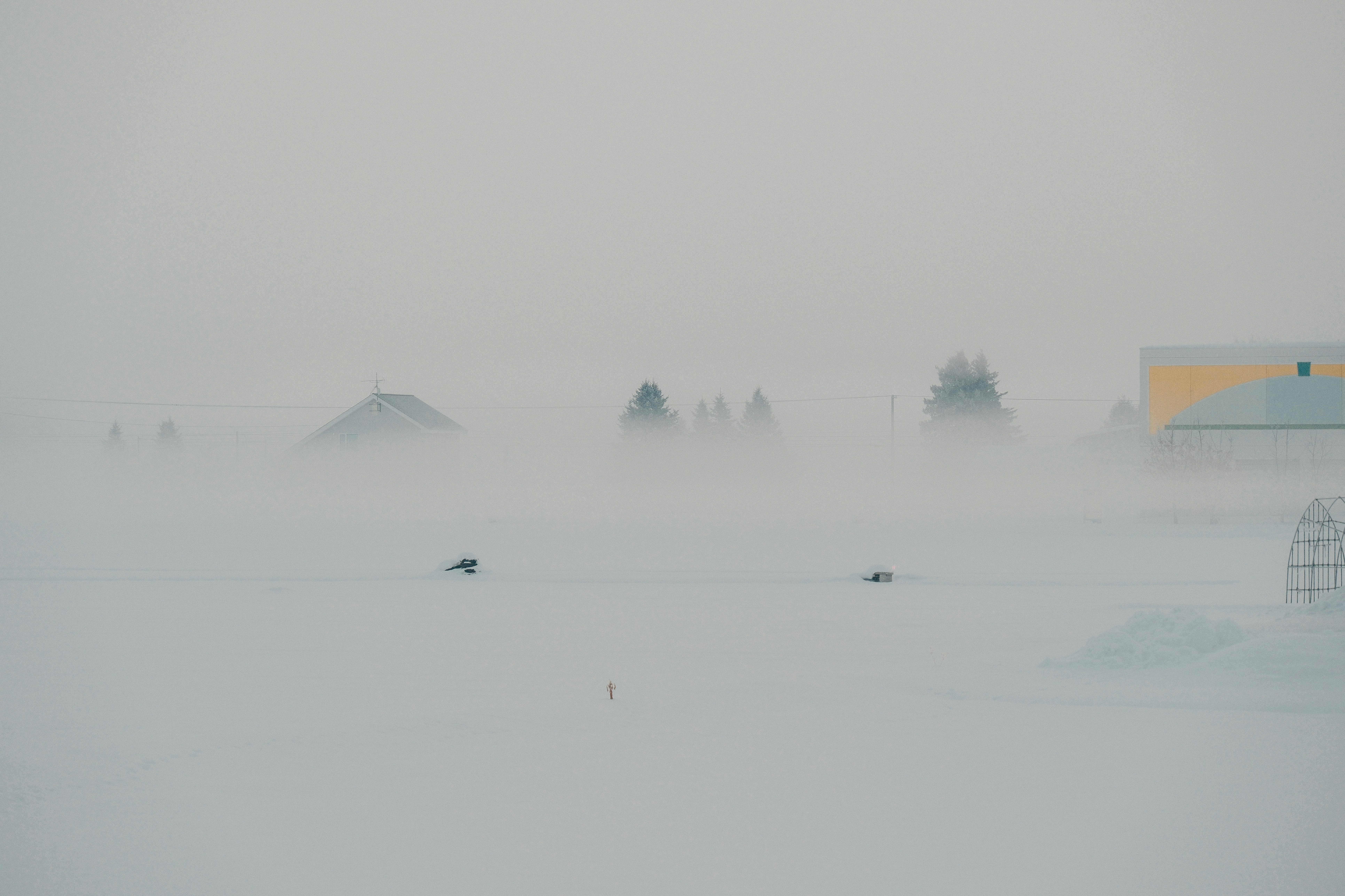 A serene winter landscape shrouded in soft fog, revealing silhouettes of distant structures and trees. The muted tones create a tranquil, ethereal atmosphere.
