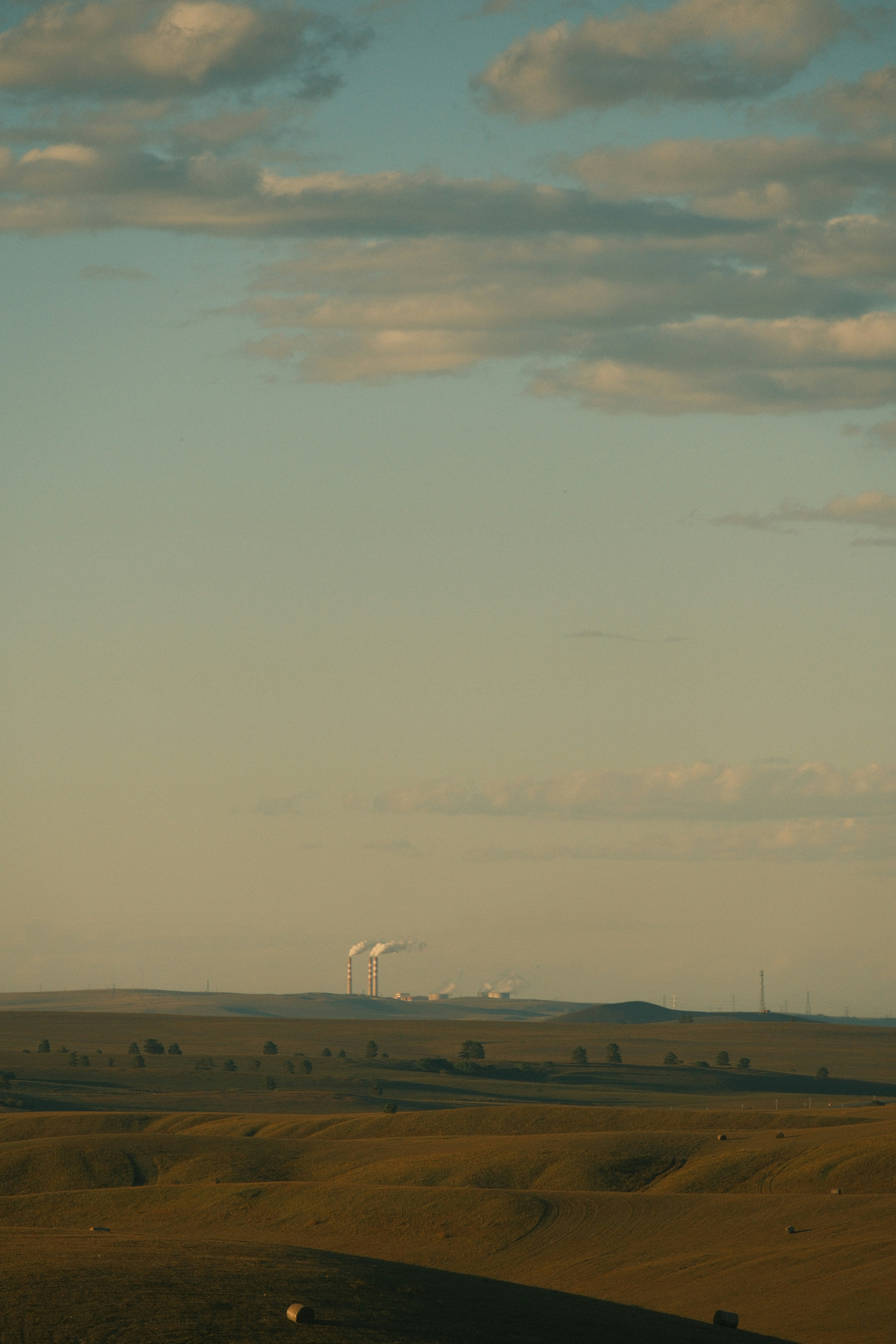 Distant power plant smokestacks rise above rolling hills under a soft sky, illustrating the contrast between nature and industry.