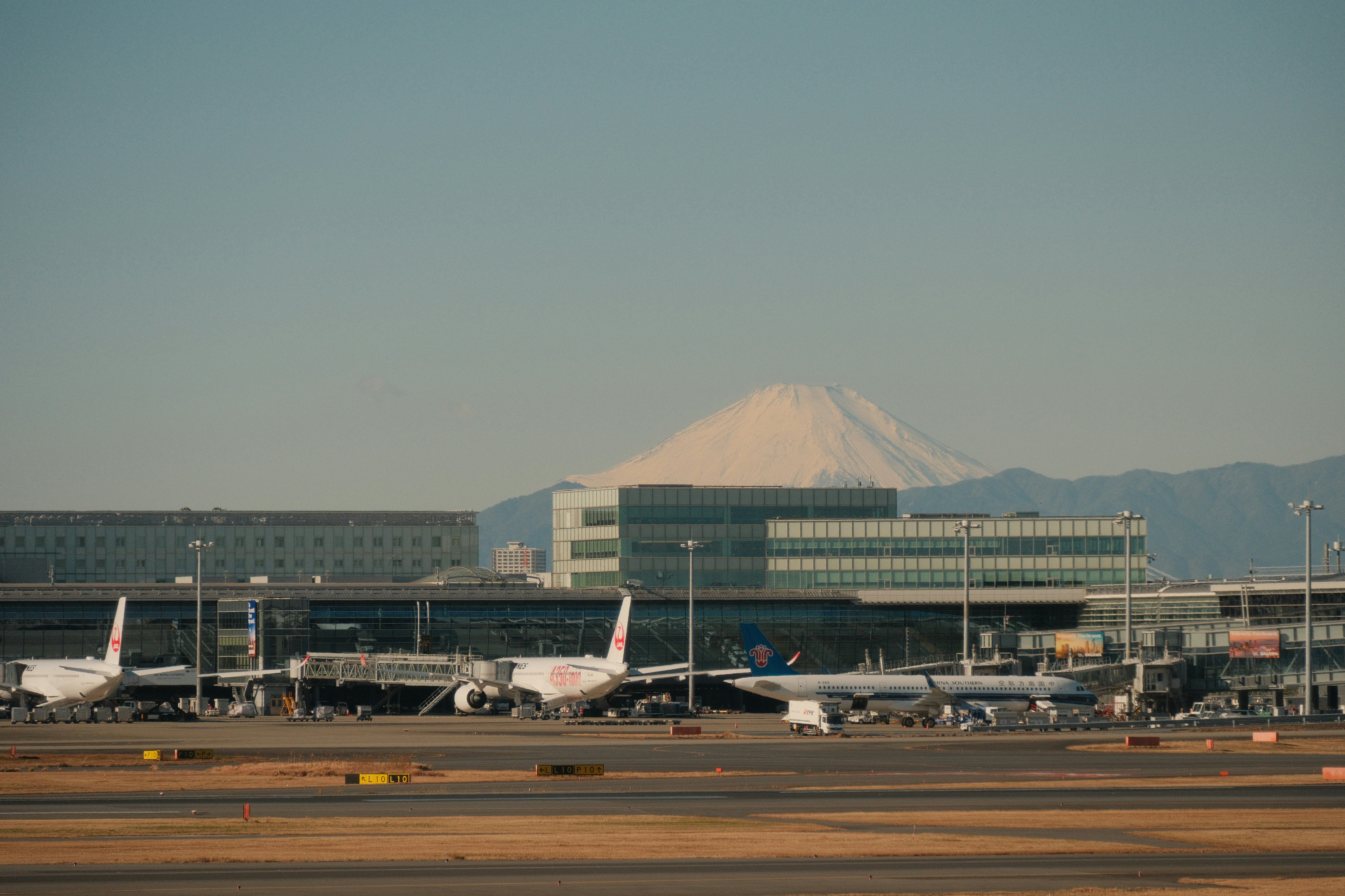 Mount Fuji rises gracefully behind a bustling airport, framed by modern architecture and aircraft on the tarmac.
