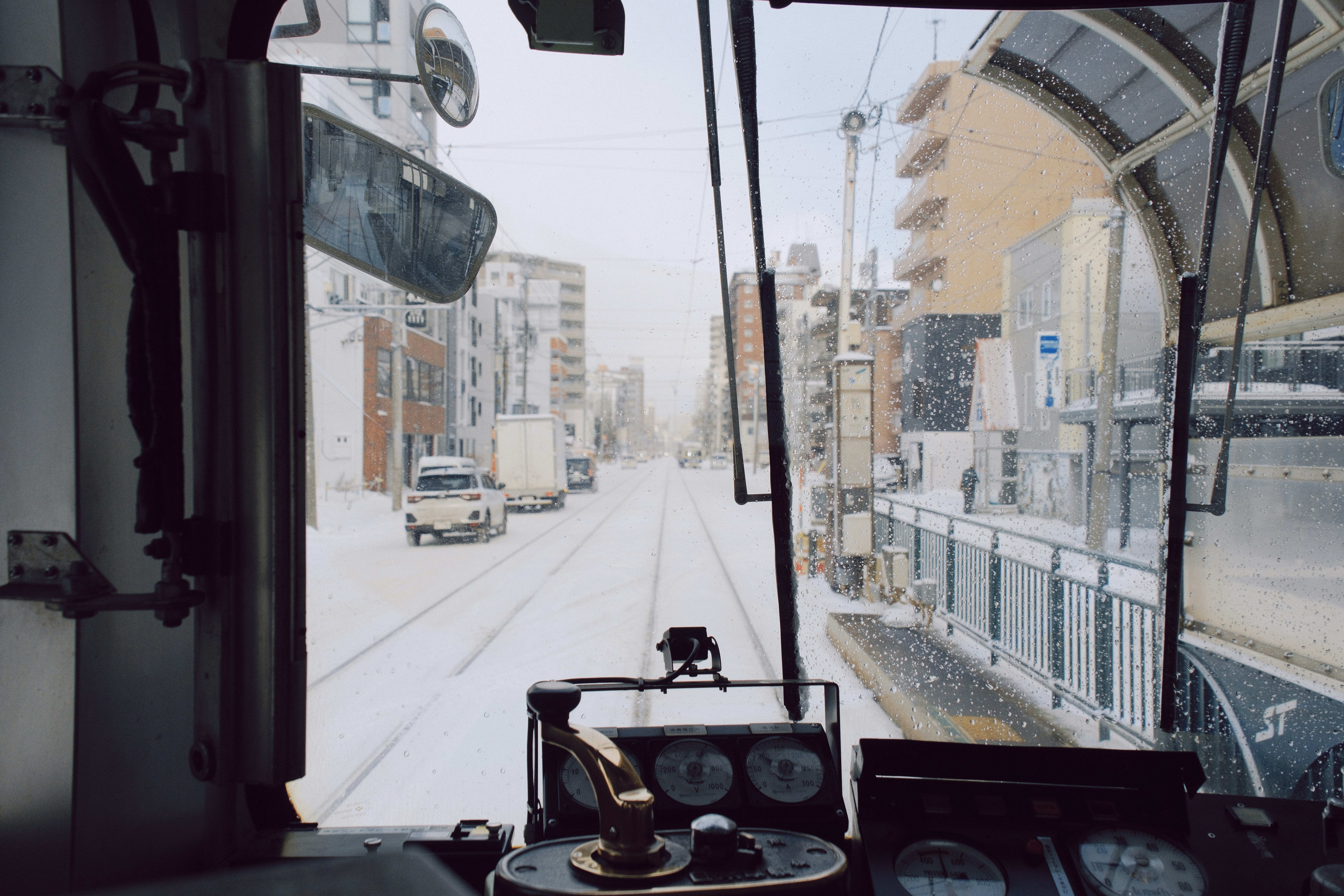 View from the driver's cabin of a snow-covered tram navigating through an urban landscape filled with softly falling snow.