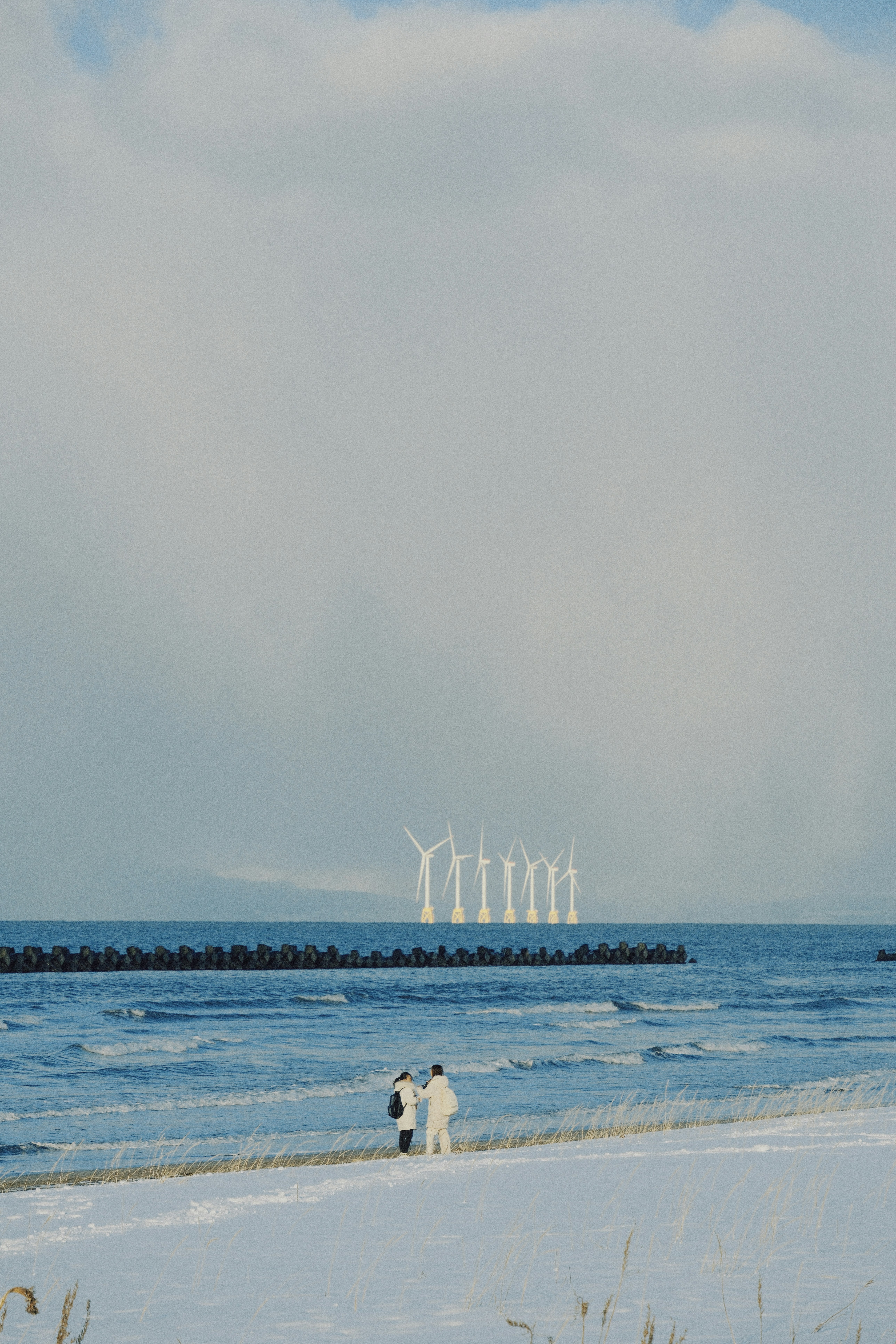 Couple walking hand in hand along a snowy beach with wind turbines in the background. The scene captures a serene winter day by the sea.