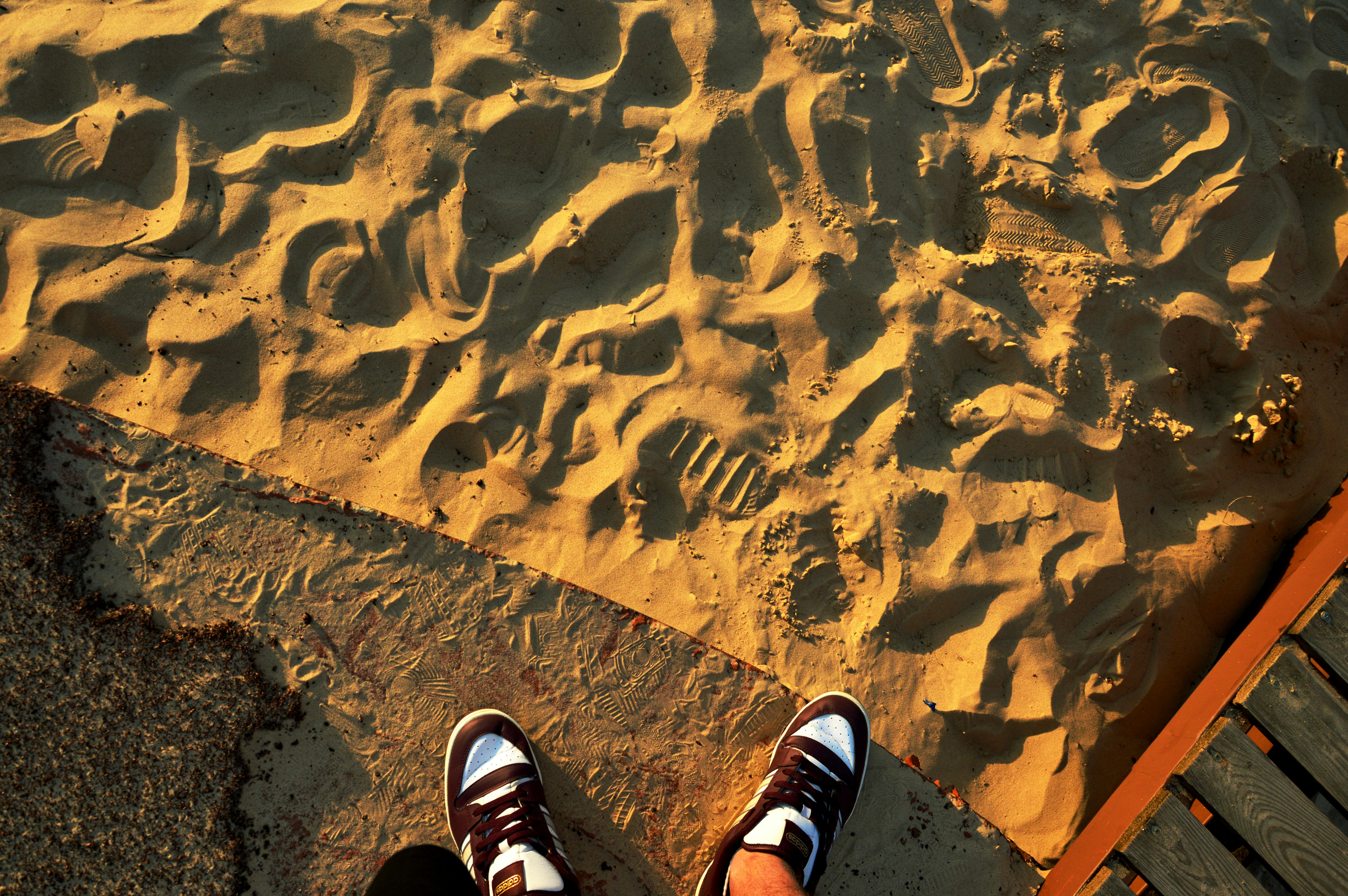 Aerial view of textured sandy surface with distinct footprints and a pair of sneakers at the edge. The warm tones of the sand contrast with the footwear.